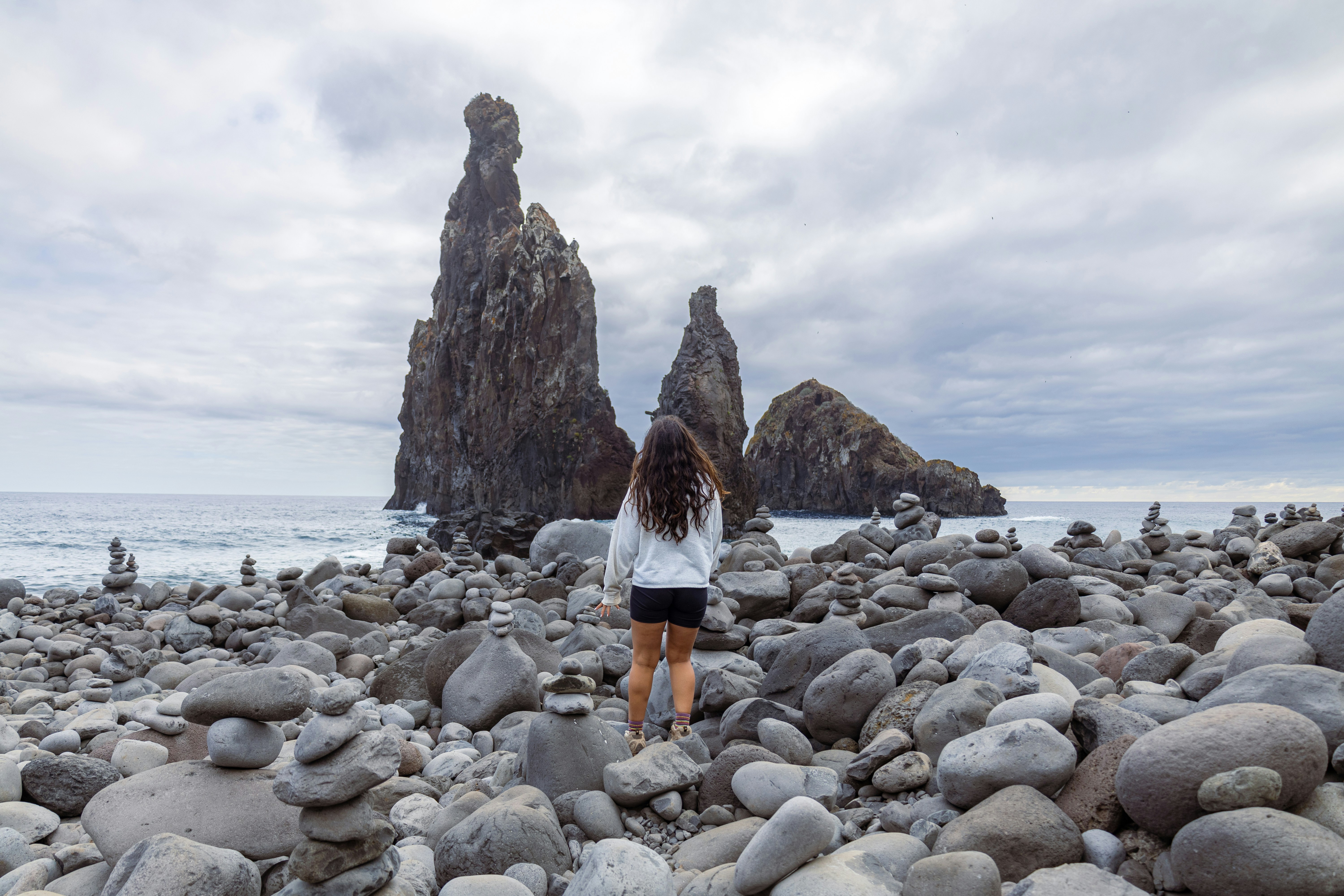 Person walking on a rocky beach with towering sea stacks under a cloudy sky.