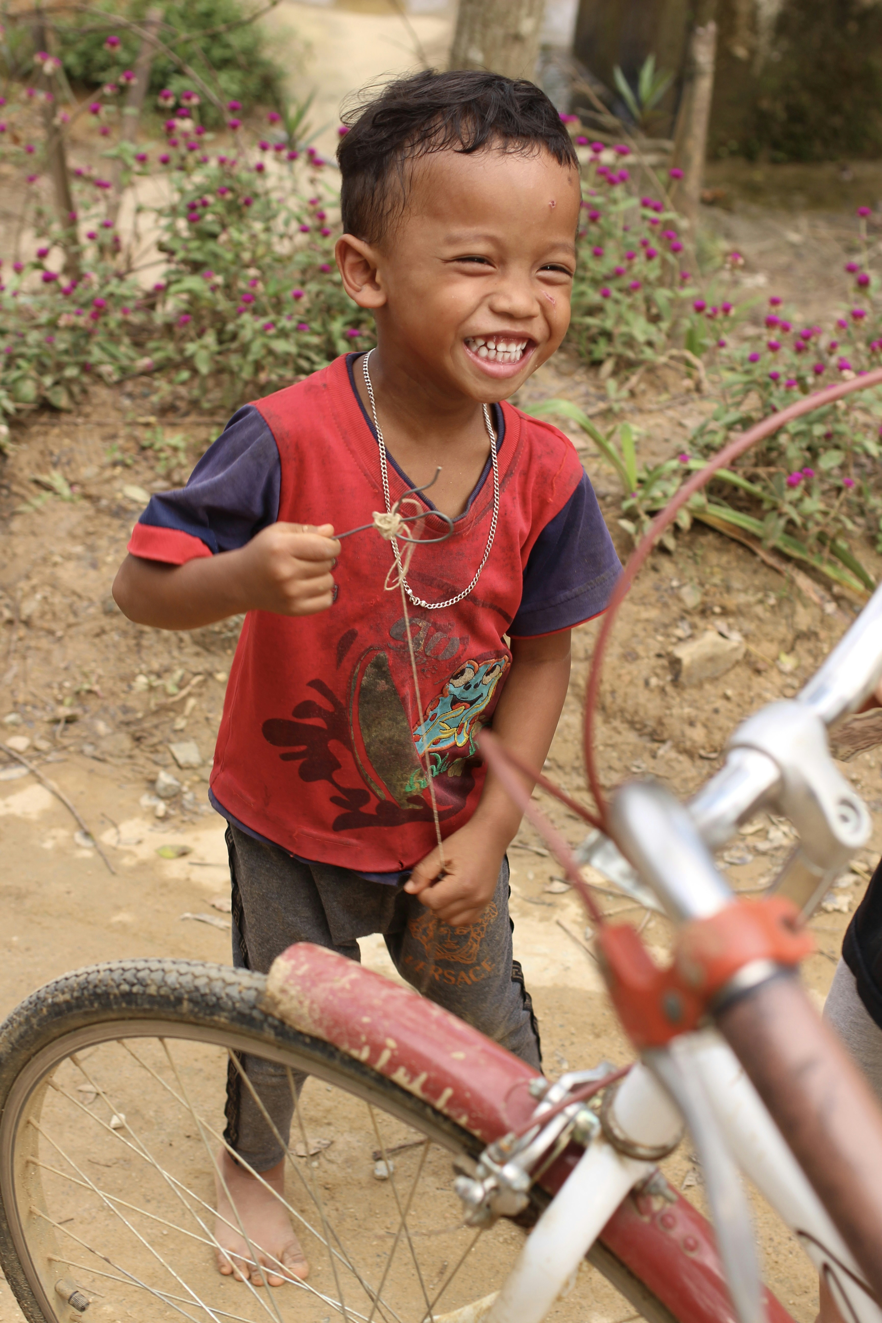 Young boy with a big smile holding a twig near his bicycle, surrounded by blooming flowers in a natural setting.