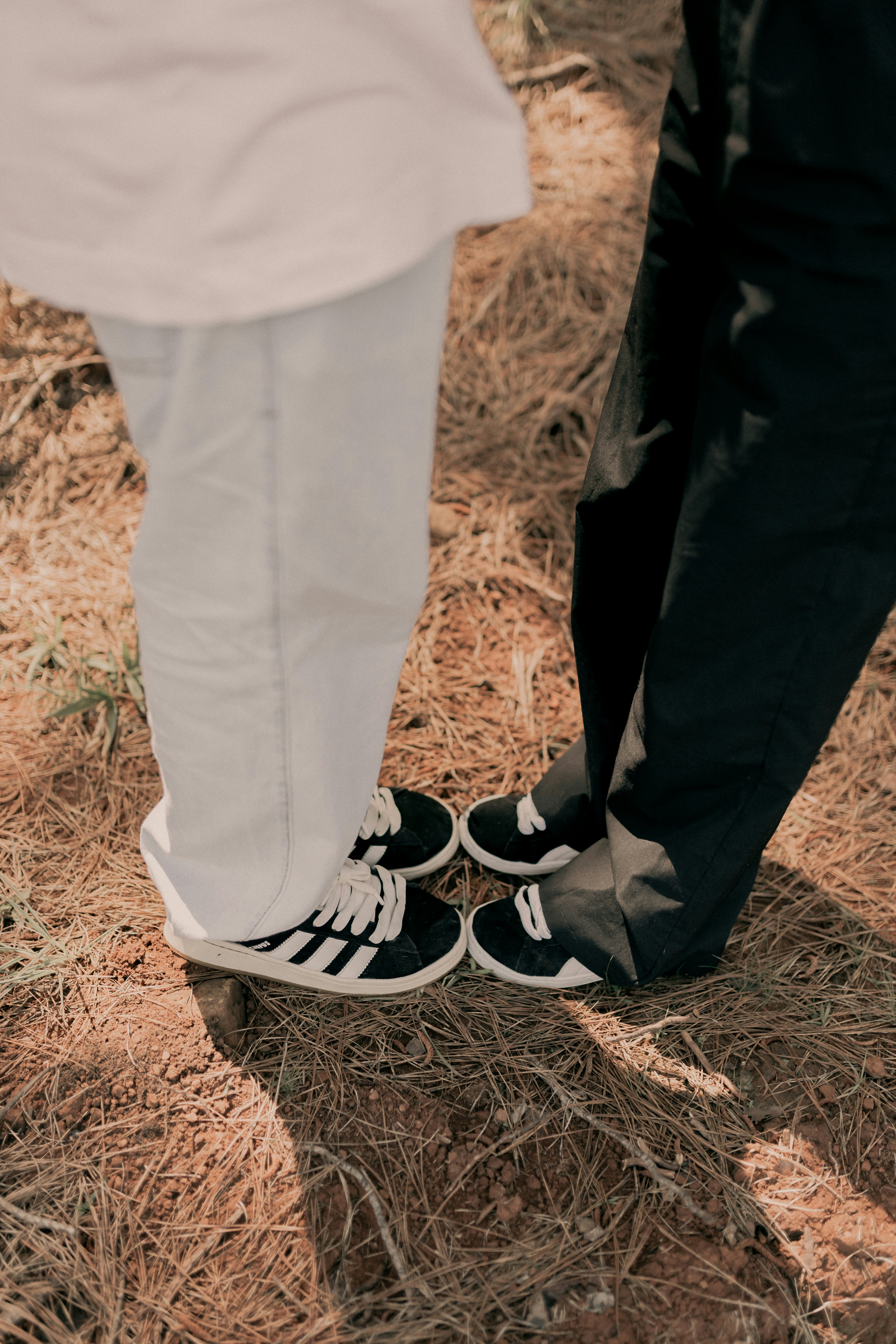 Two pairs of sneakers, one white and one black, stand closely together on a bed of pine needles, symbolizing unity and connection.