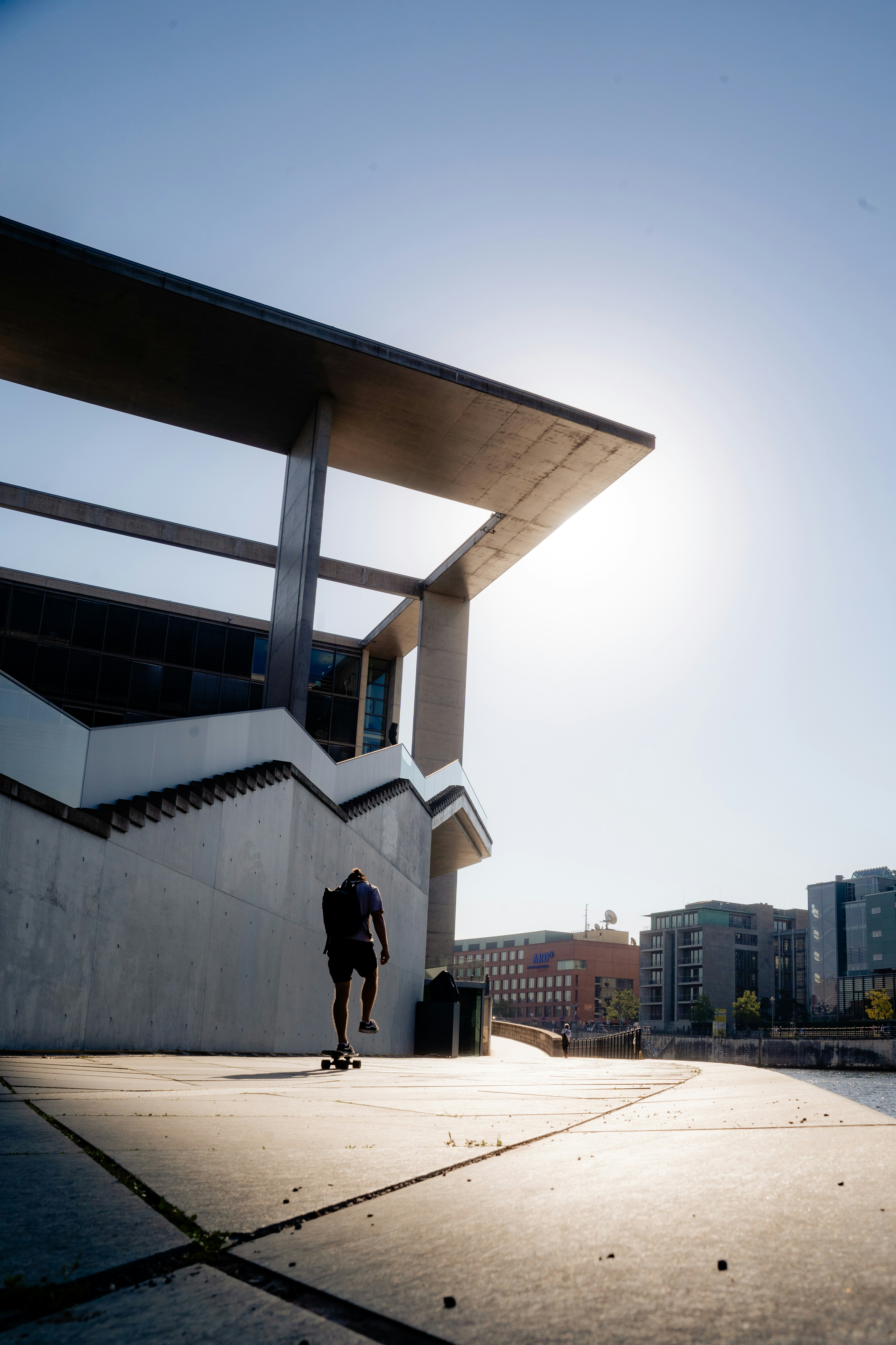 Skateboarder traversing under a modern structure with the sun casting sharp shadows on concrete, contrasting with the soft morning light.