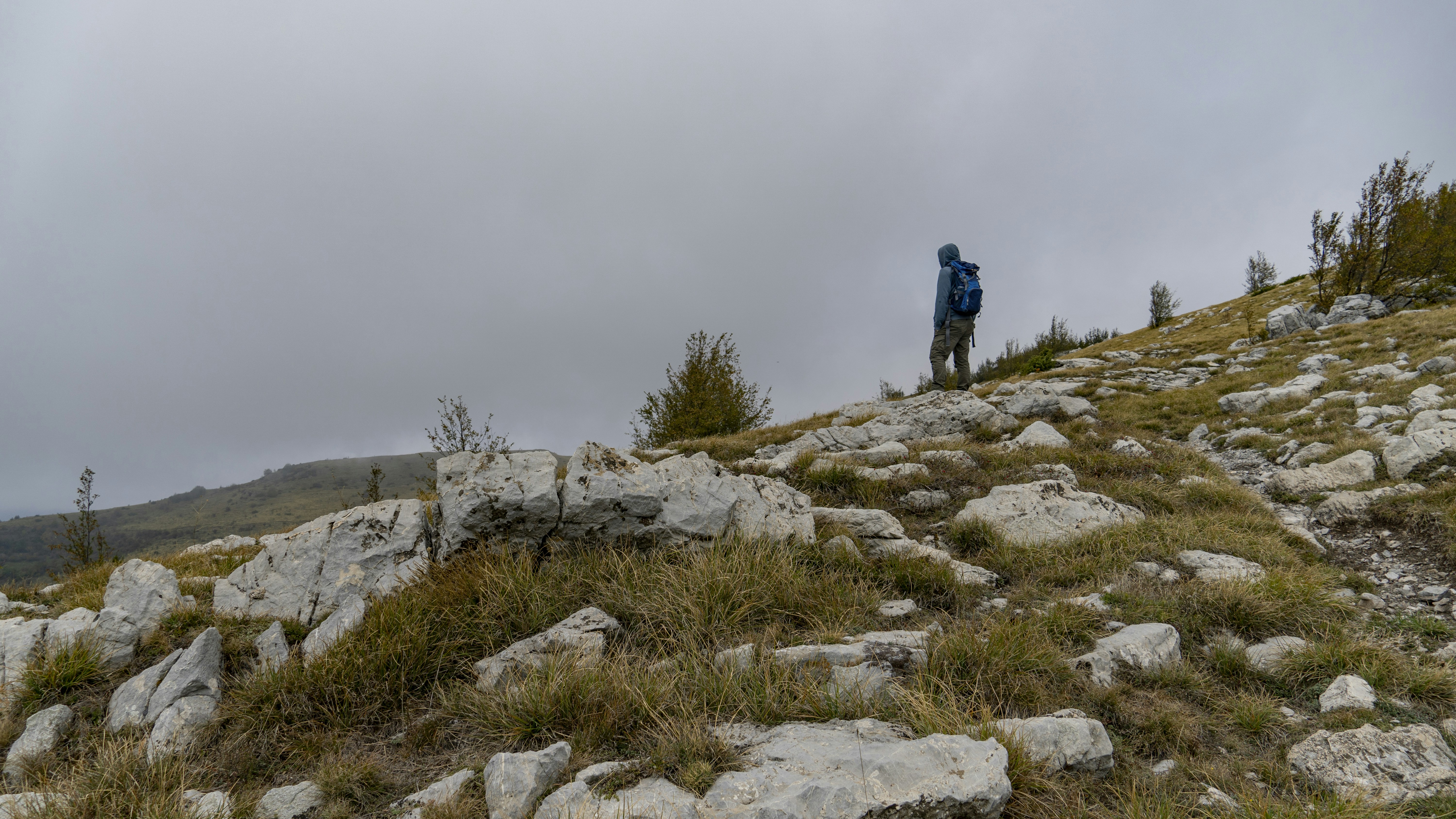 Hiker navigating rocky terrain on a foggy hillside, surrounded by sparse vegetation. The atmosphere conveys a sense of adventure and solitude.