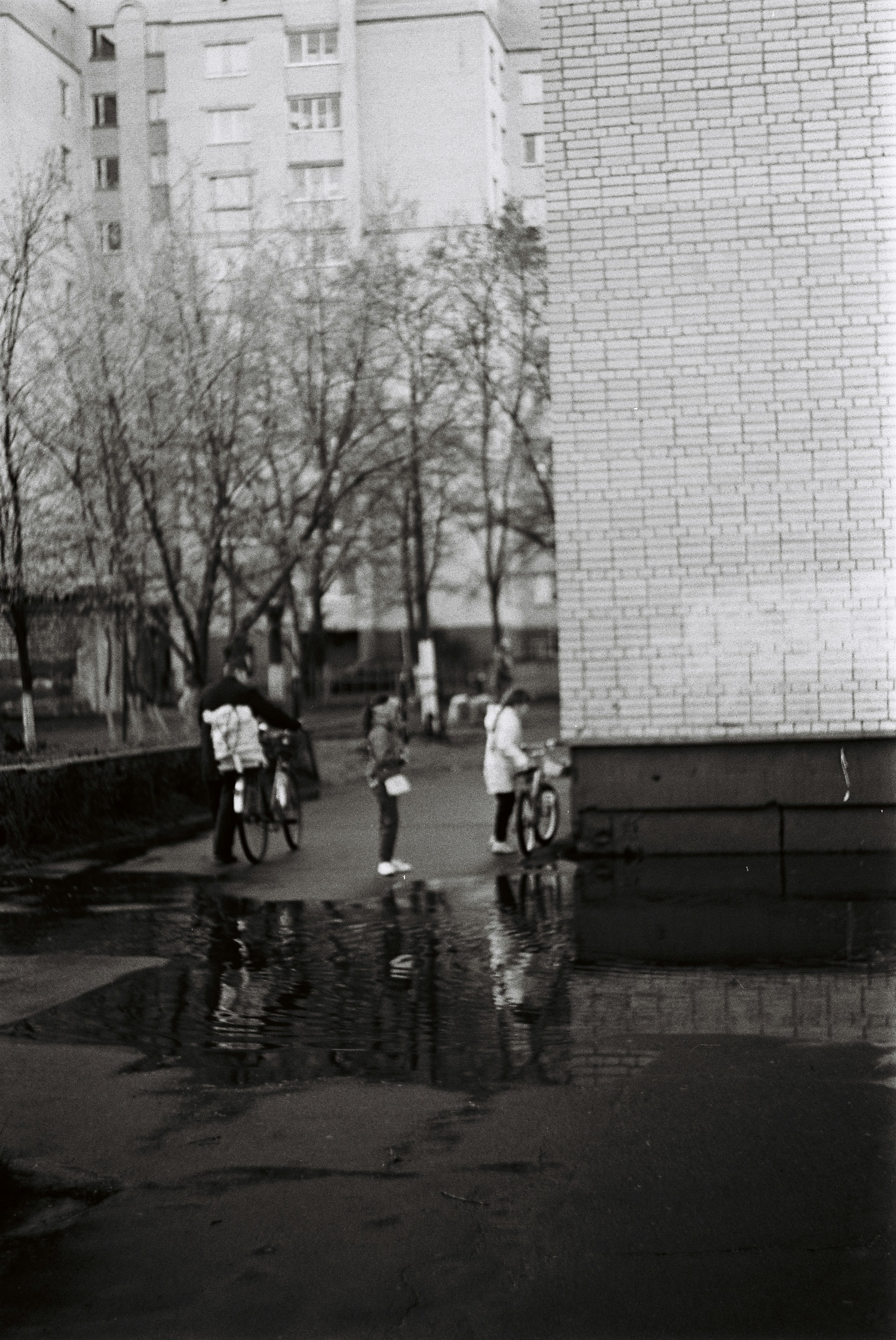 Black-and-white street photograph capturing children and cyclists near a brick building, mirrored by a rain-filled puddle.