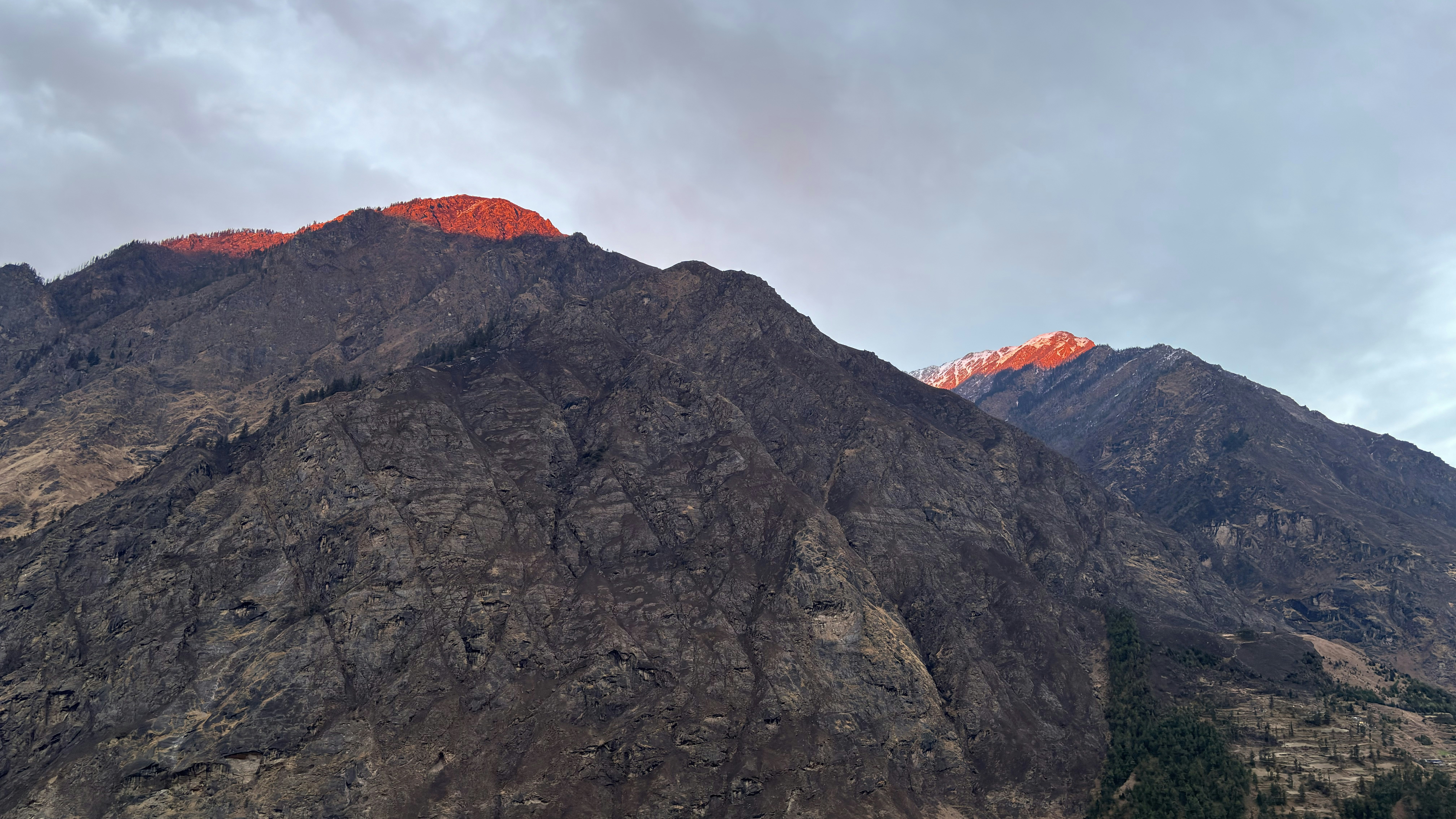 Mountain peaks illuminated by the orange glow of a setting sun beneath a cloudy sky.