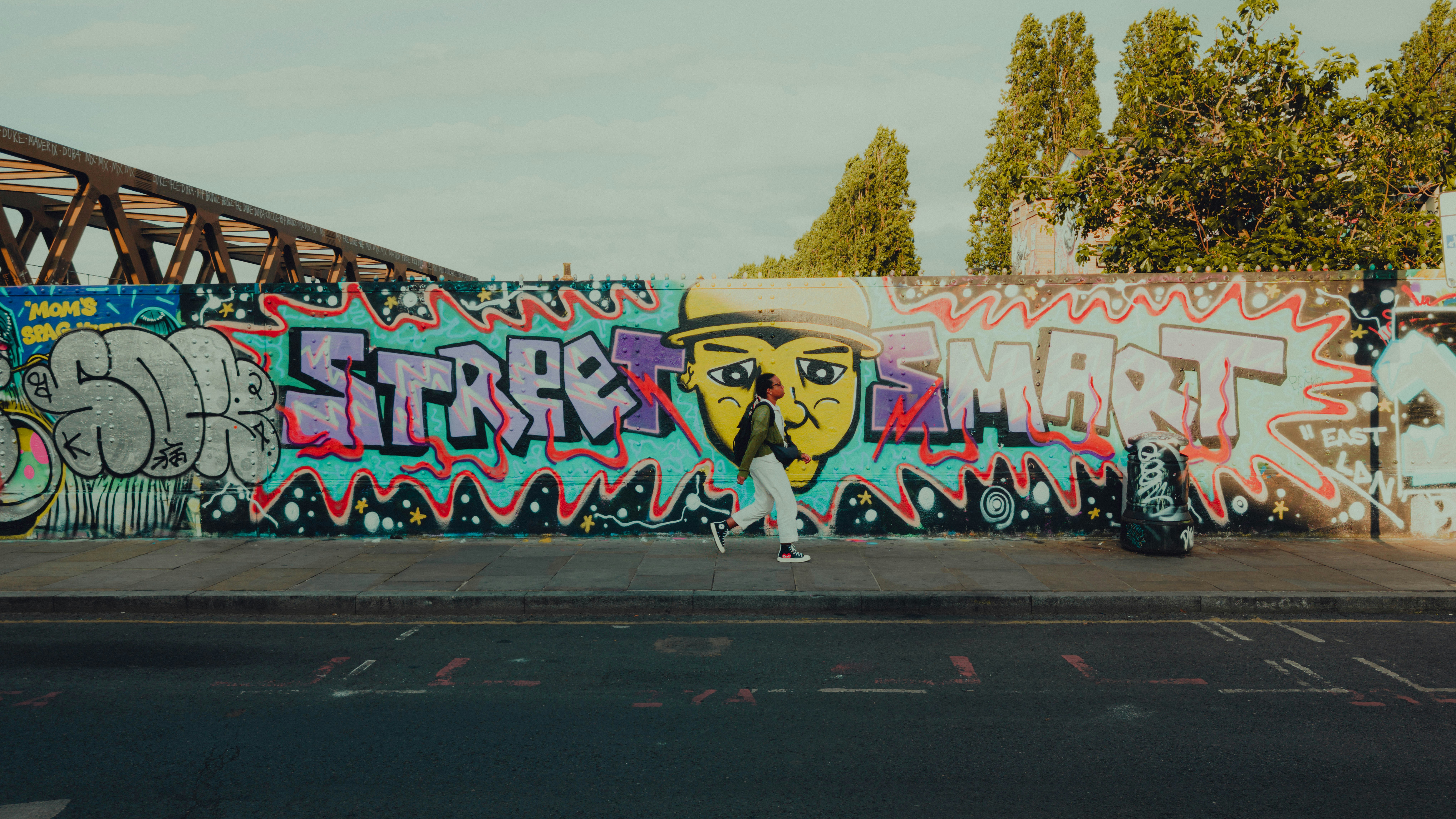 A passerby walks along a graffiti-covered wall dominated by a large yellow-faced mural. The scene captures street art and everyday movement in a vibrant urban setting.