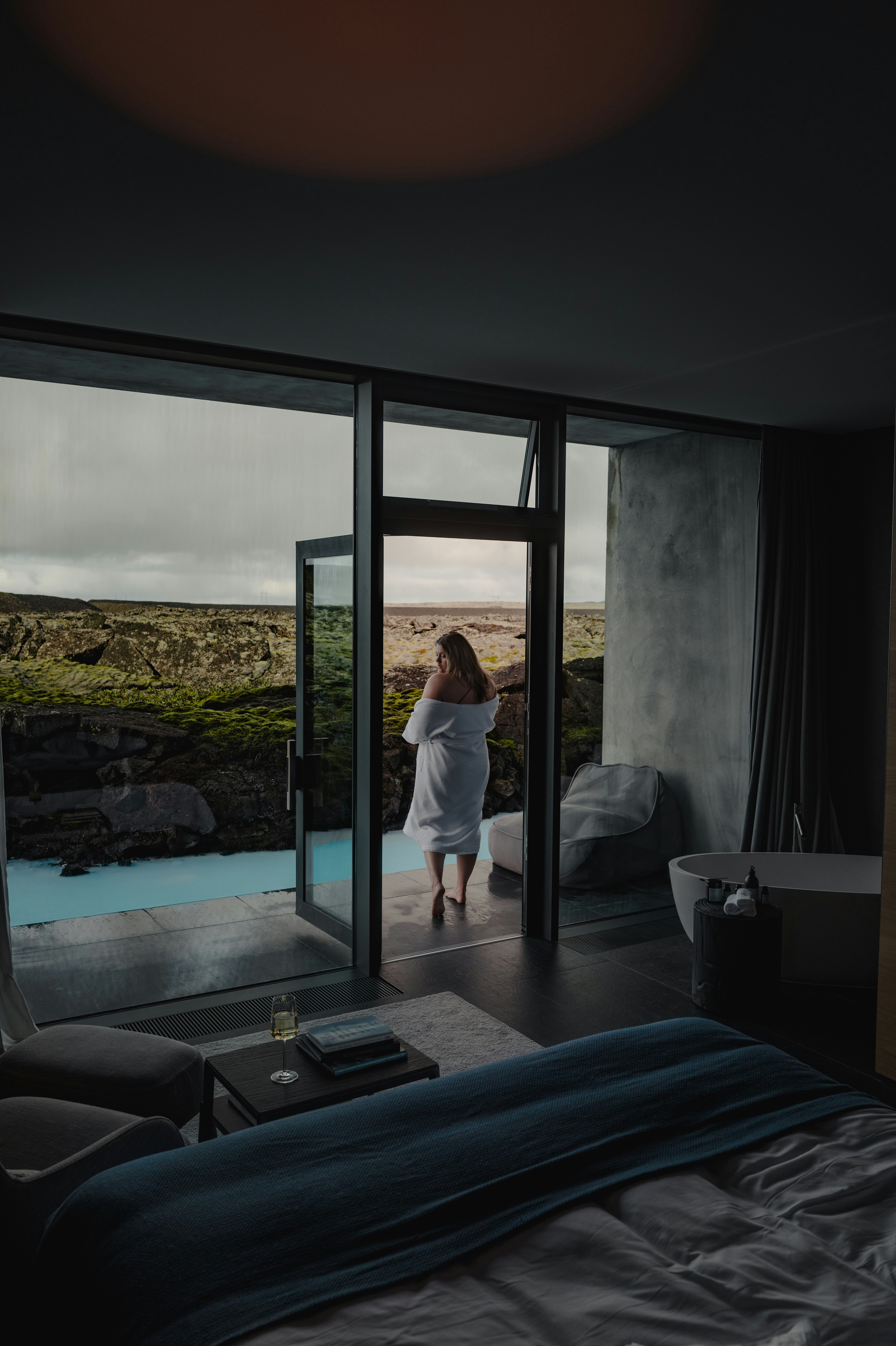 Person in a bathrobe standing by an open door, overlooking a tranquil outdoor pool and rocky landscape.
