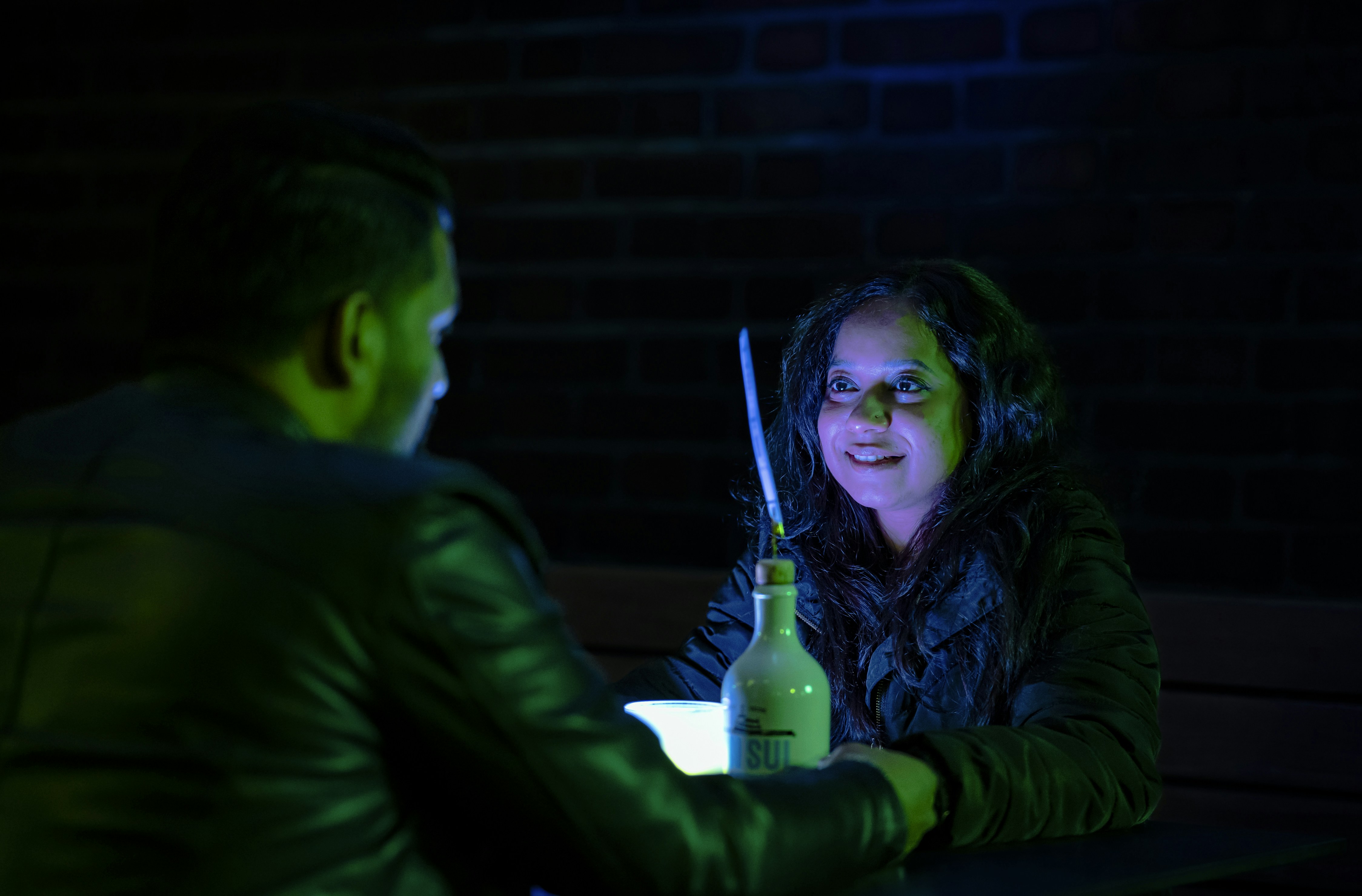 Two people converse under moody neon lighting, highlighting their expressions against a brick wall.