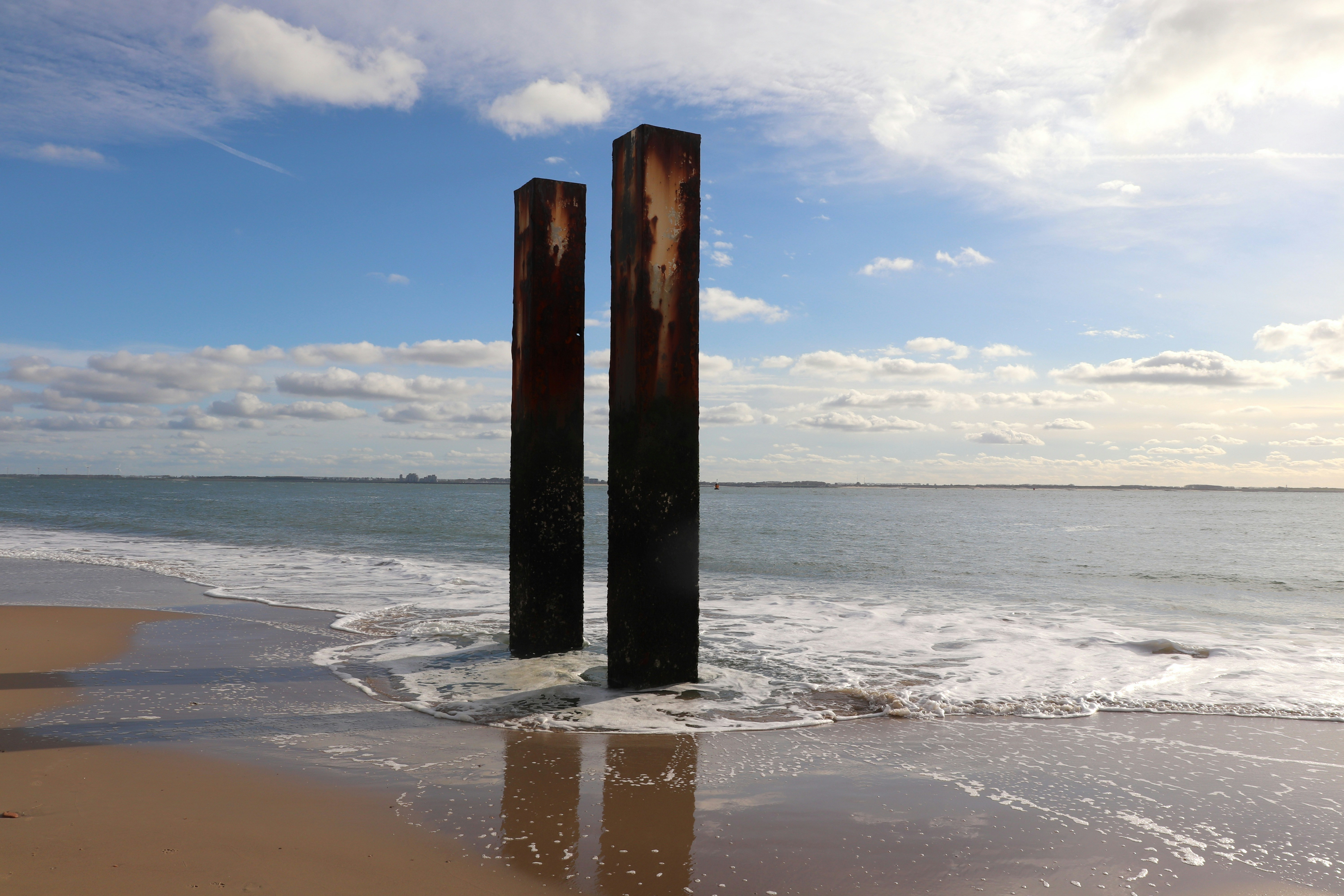 Two rusted vertical pilings rise from the wet sand at the edge of a calm sea, captured in this photograph. The scene highlights weathered texture and a serene maritime mood.