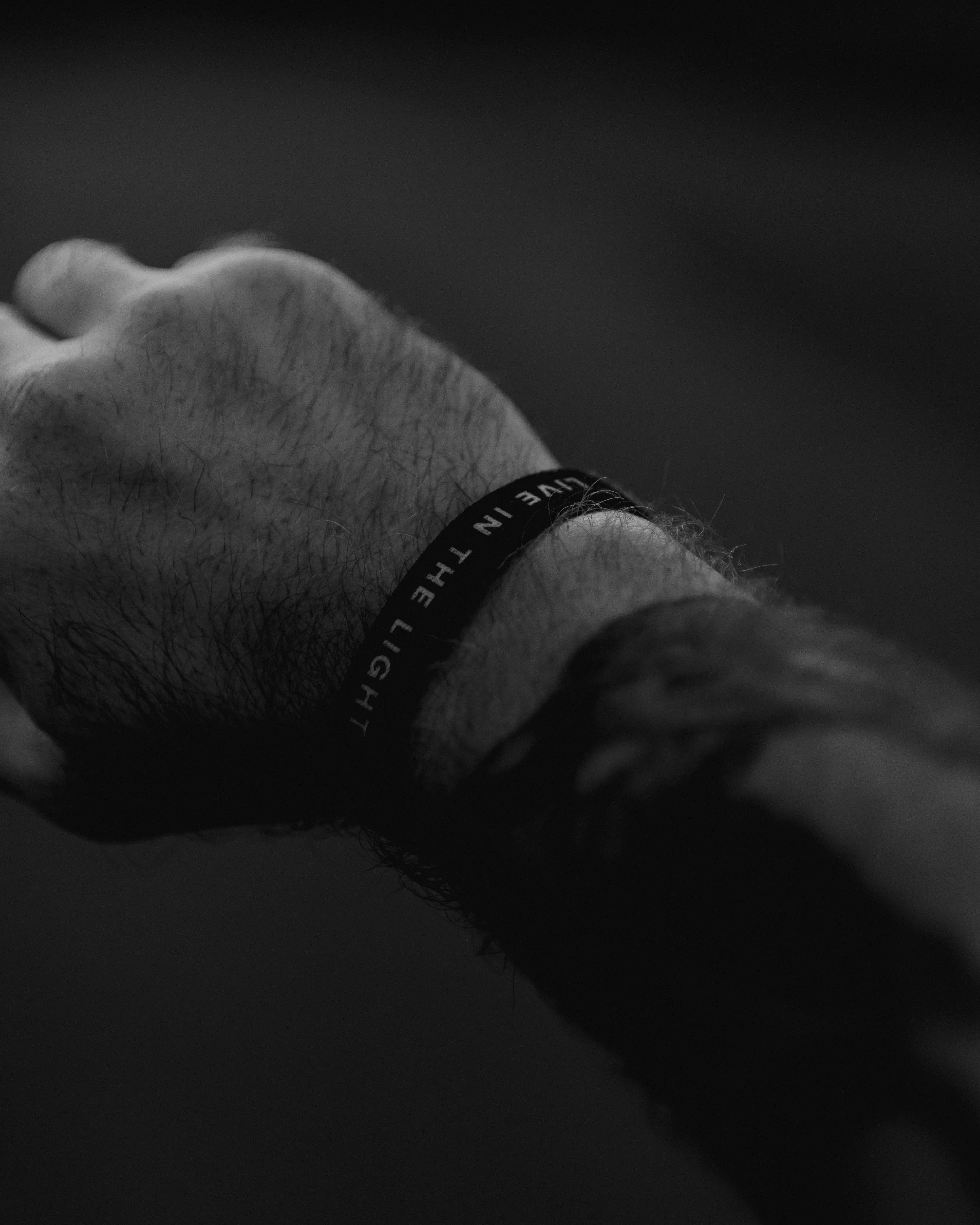 Close-up of a hand wearing a black wristband with the phrase 'LIVE IN THE LIGHT' prominently displayed. The monochrome tones emphasize the message's significance.