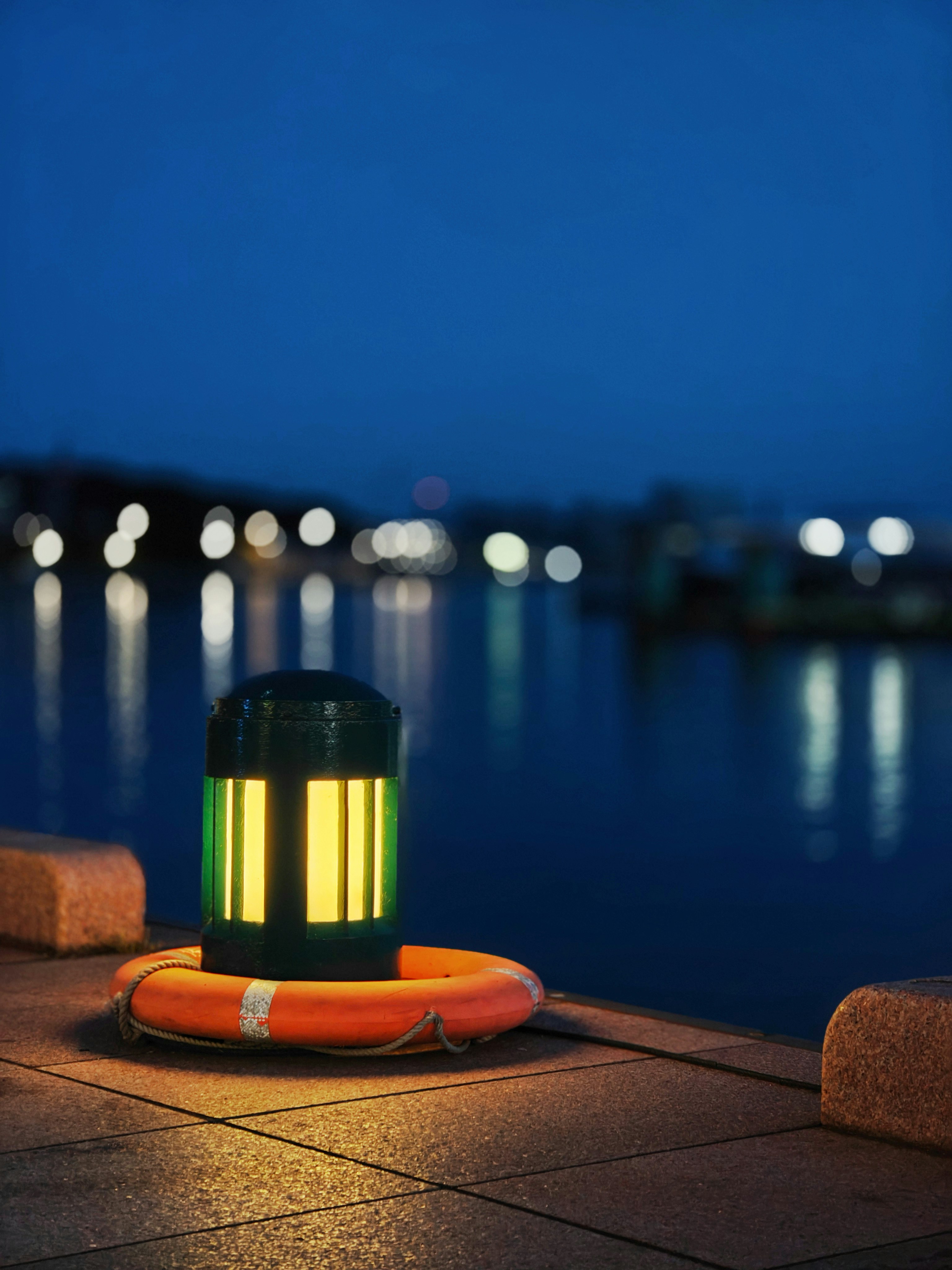 Green cylindrical lantern on a quay glows yellow-green, casting reflections on the wet tiles while blurred harbor lights shimmer in the blue evening water.