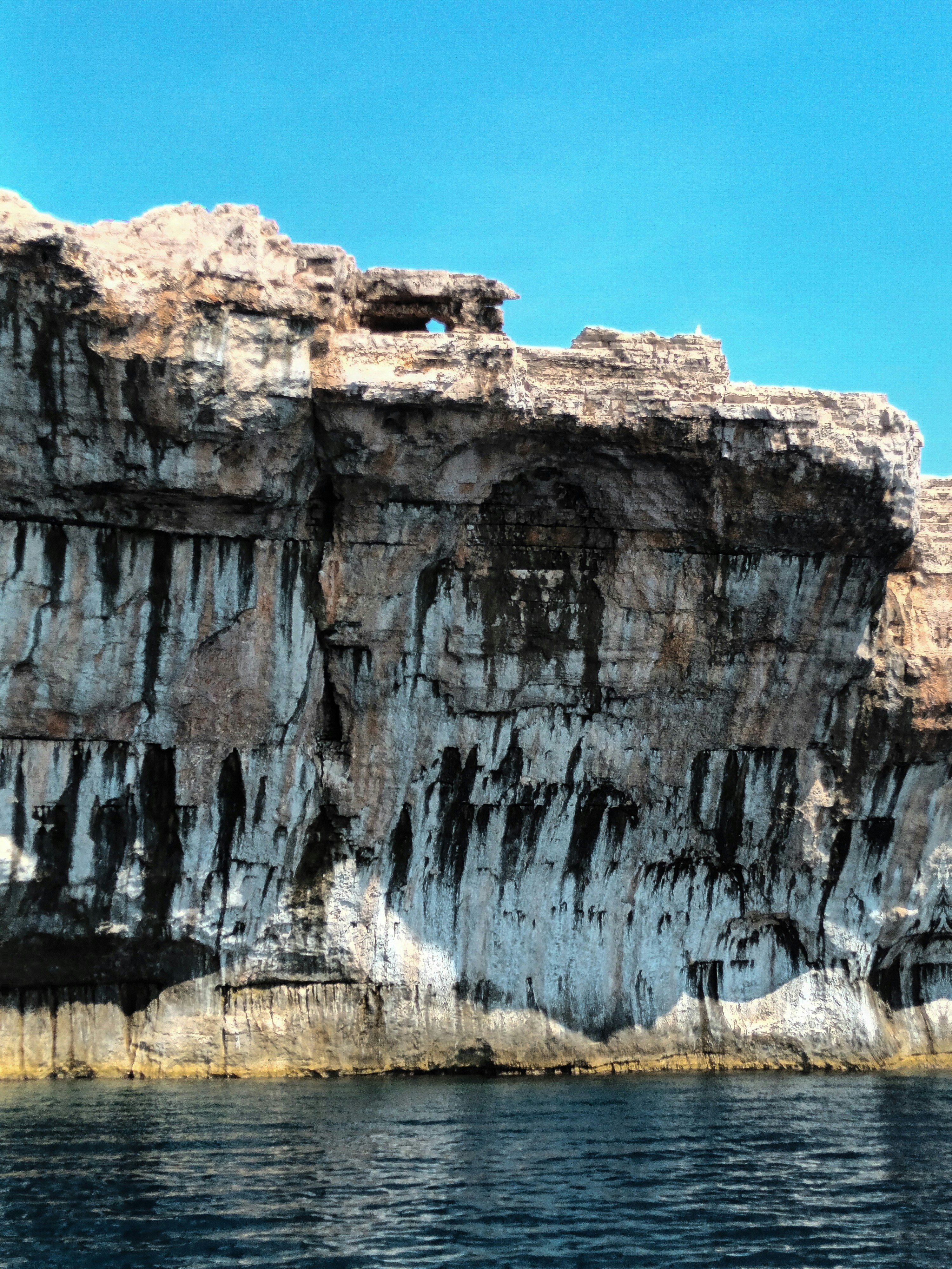 Craggy limestone cliff meeting calm sea, with a small natural opening high in the rock; rugged texture and clear blue sky form a stark seascape.