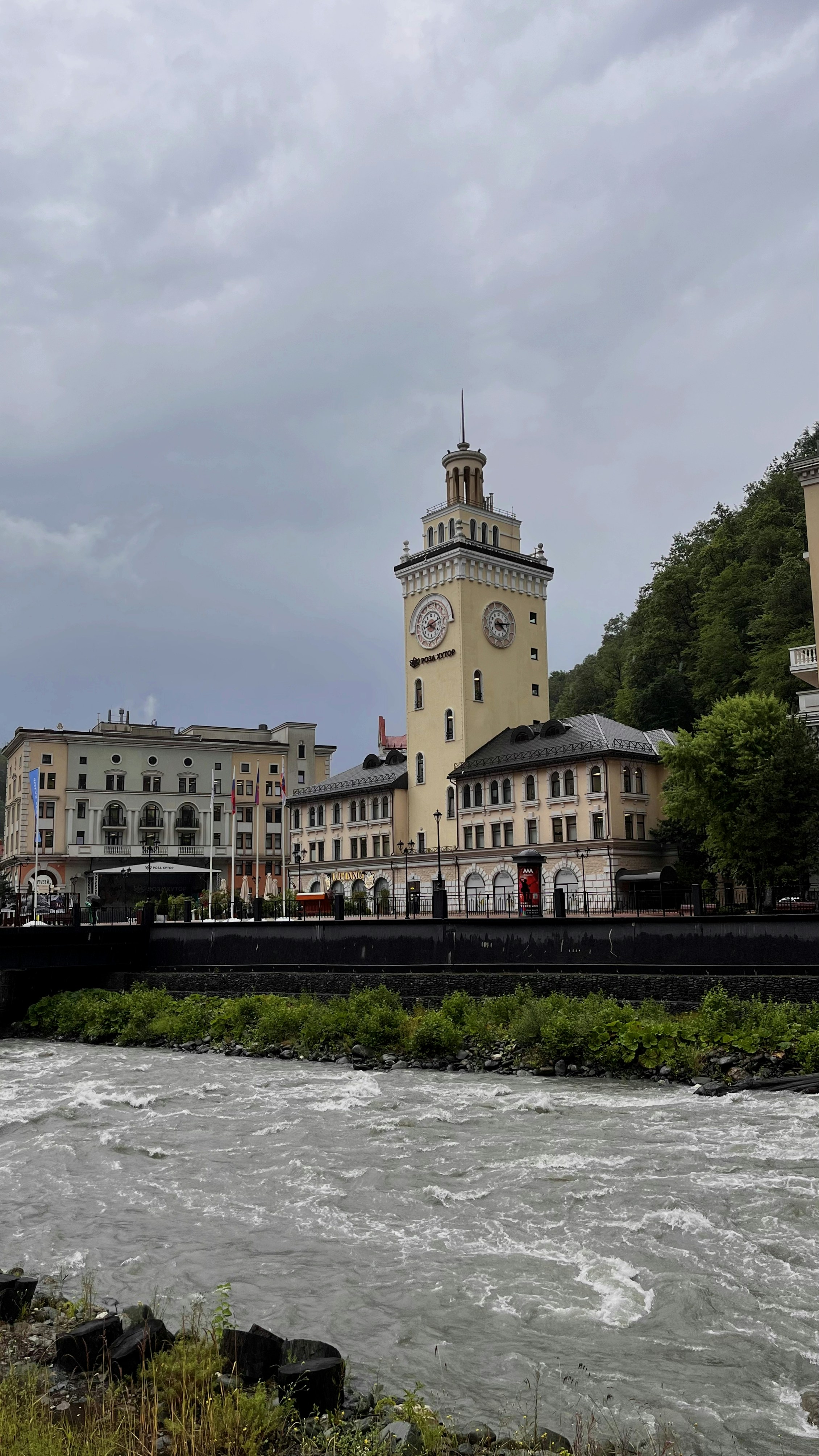 Historic clock tower overlooking a rushing river, framed by charming buildings and lush greenery. A scene that embodies the essence of a serene town.
