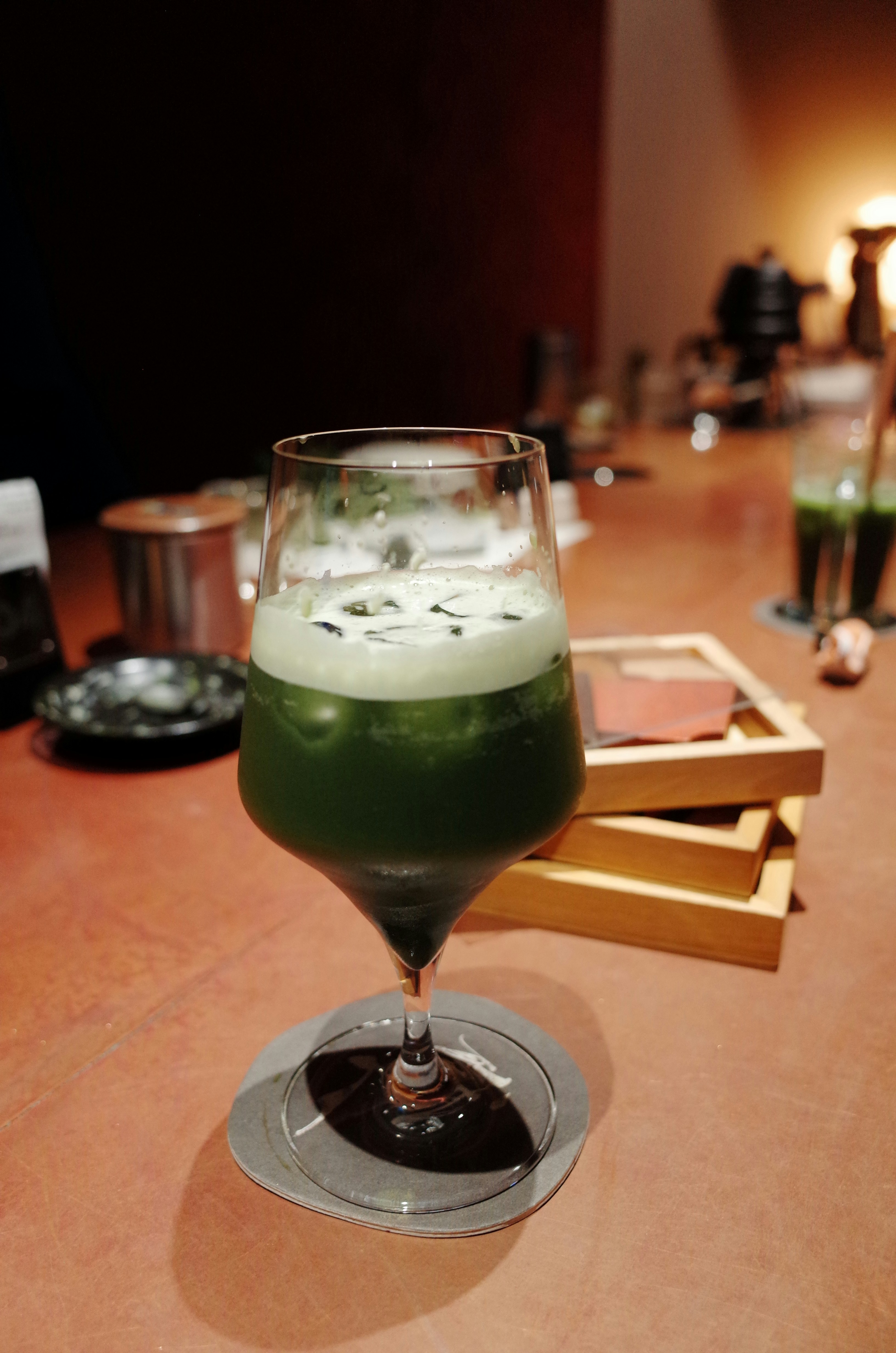 An emerald-green cocktail with a foamy top sits on a wooden bar counter, ice visible through the glass. The shallow depth of field blurs the background, drawing attention to the drink.