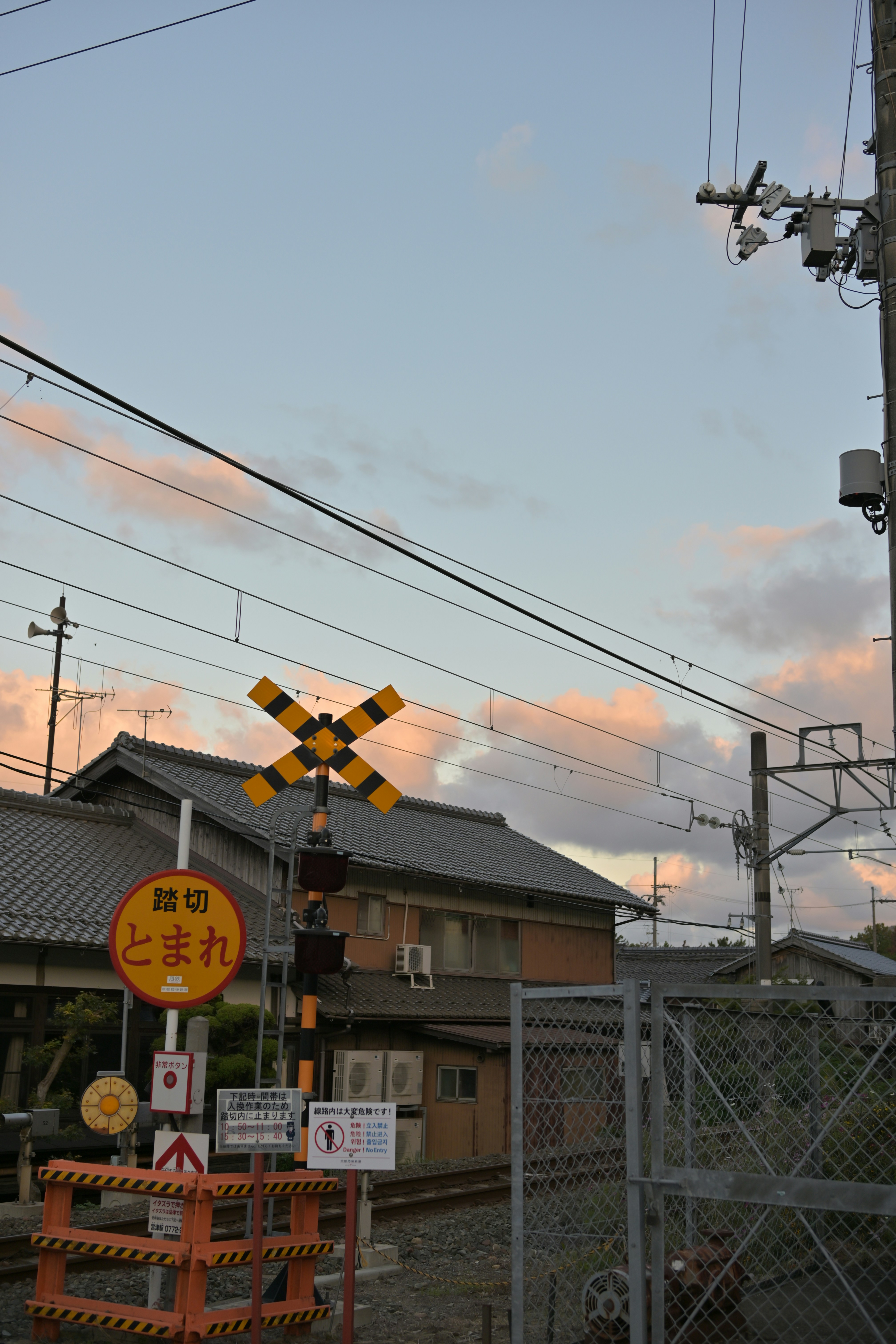 A quiet Japanese railroad crossing at dusk, featuring a yellow-and-black crossbuck and an orange circular stop sign amid residential buildings.
