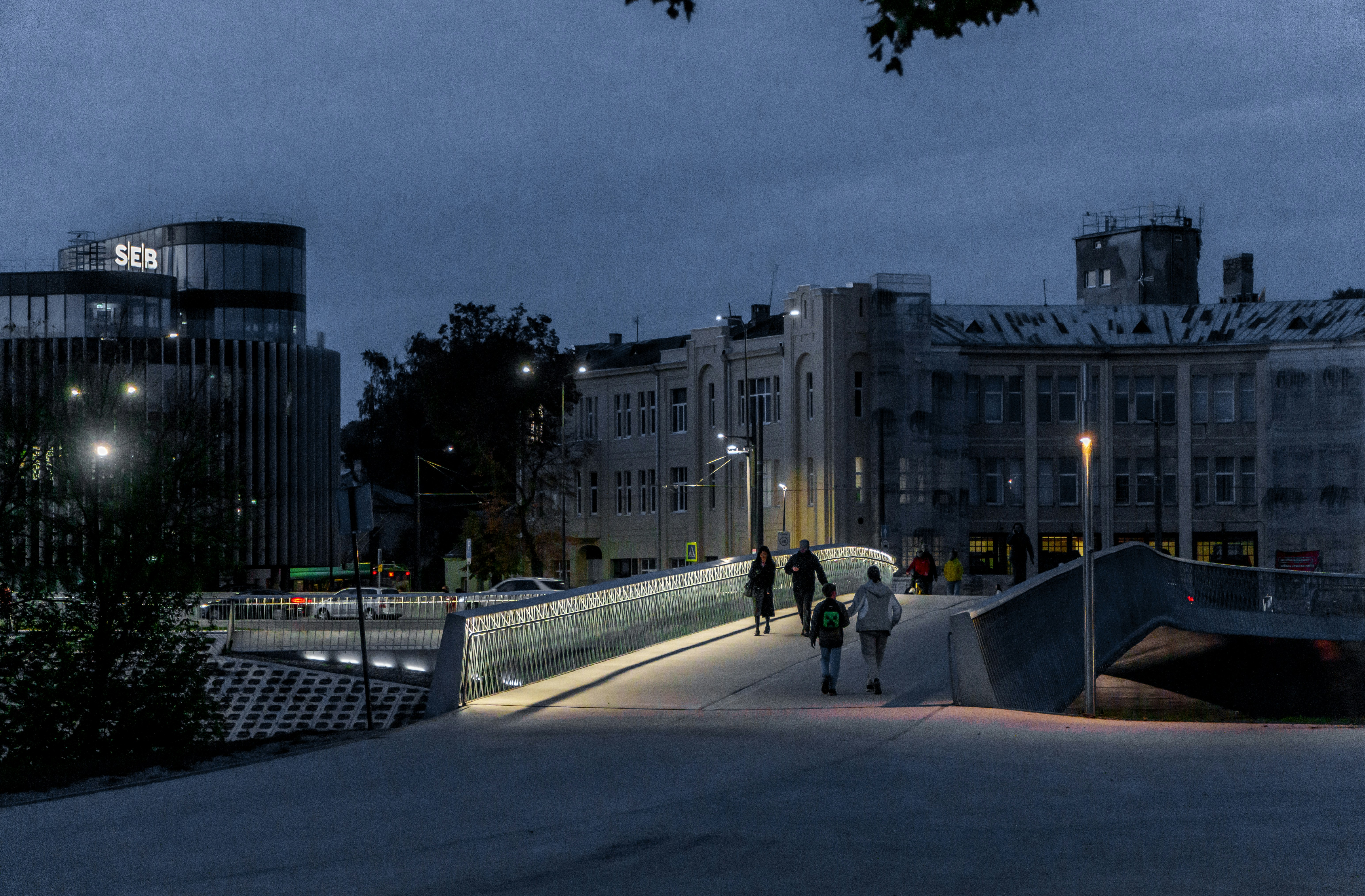 People walking across a brightly lit bridge against a city skyline at dusk.