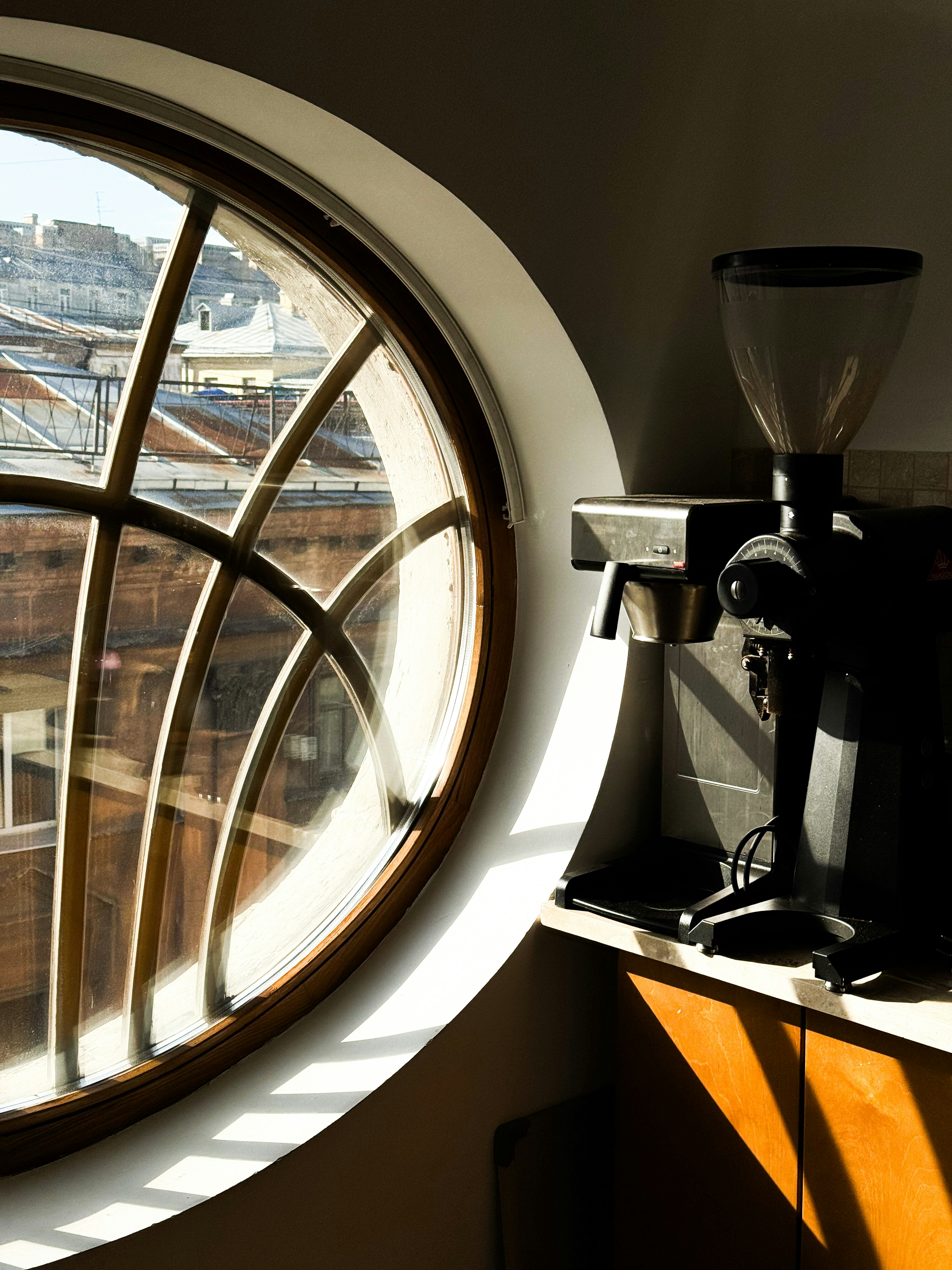 Morning light floods a round window, casting geometric shadows on a coffee grinder perched beside a wooden counter. Urban rooftops peek through the glass, while warm interior light defines texture and form.