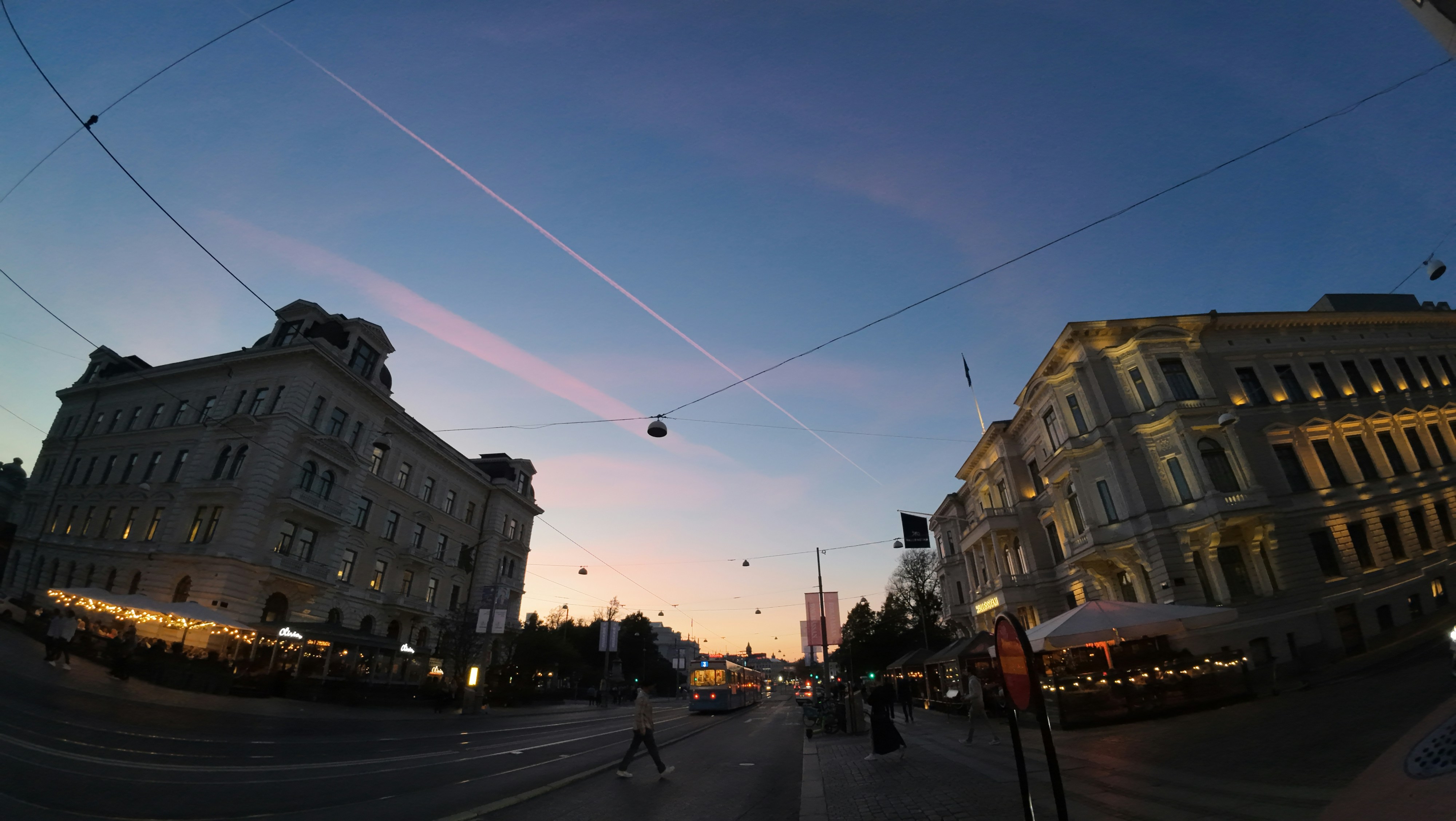 Wide-angle street scene at dusk shows tram wires crisscrossing the sky above historic buildings along a curved boulevard. The photo captures motion and ambient city lights with an expansive, fisheye perspective.