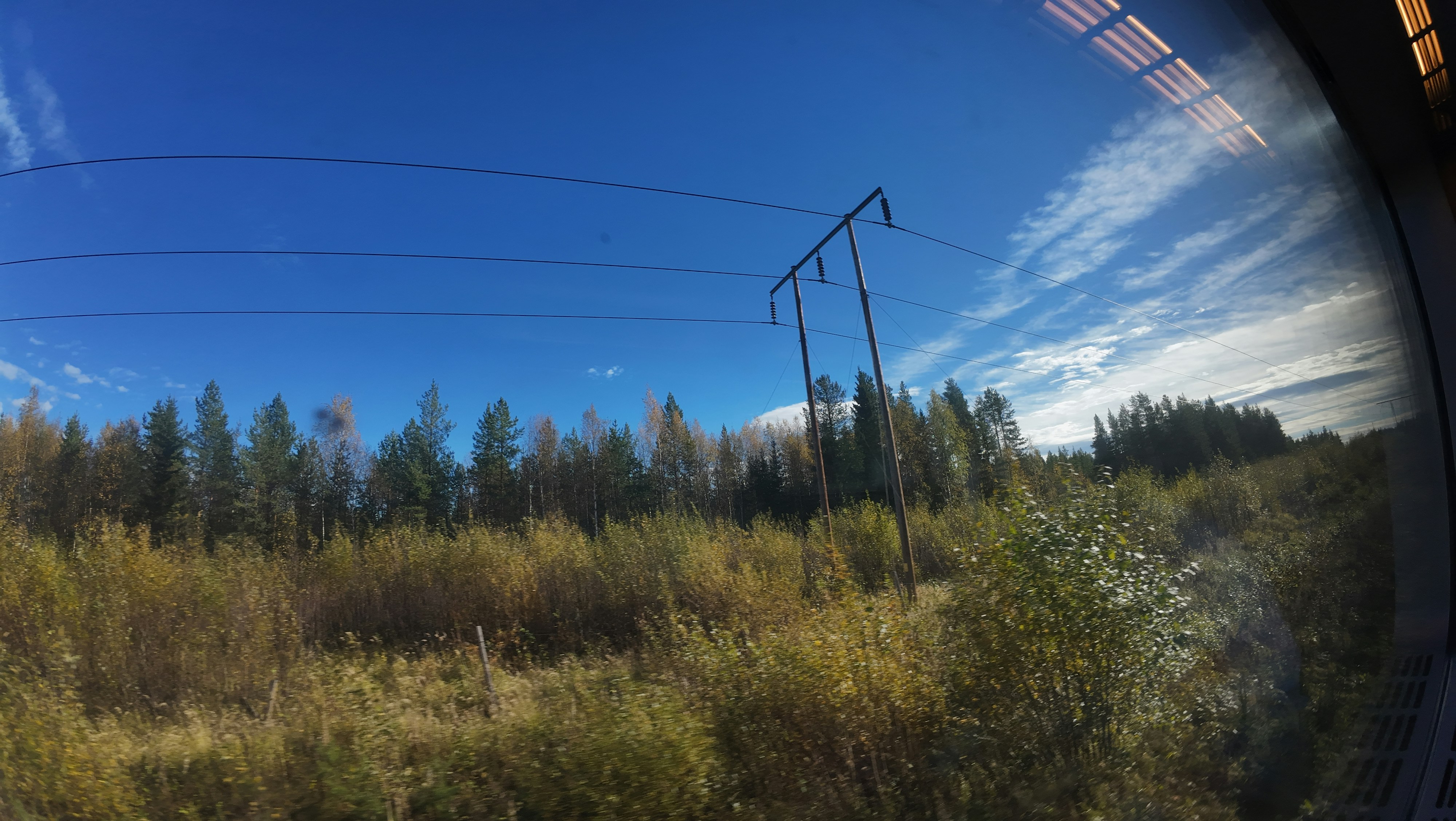 Wide-angle landscape of a golden prairie with power lines crossing the scene beneath a bright blue sky. The curved perspective suggests a panoramic view with a window-like distortion along the horizon.