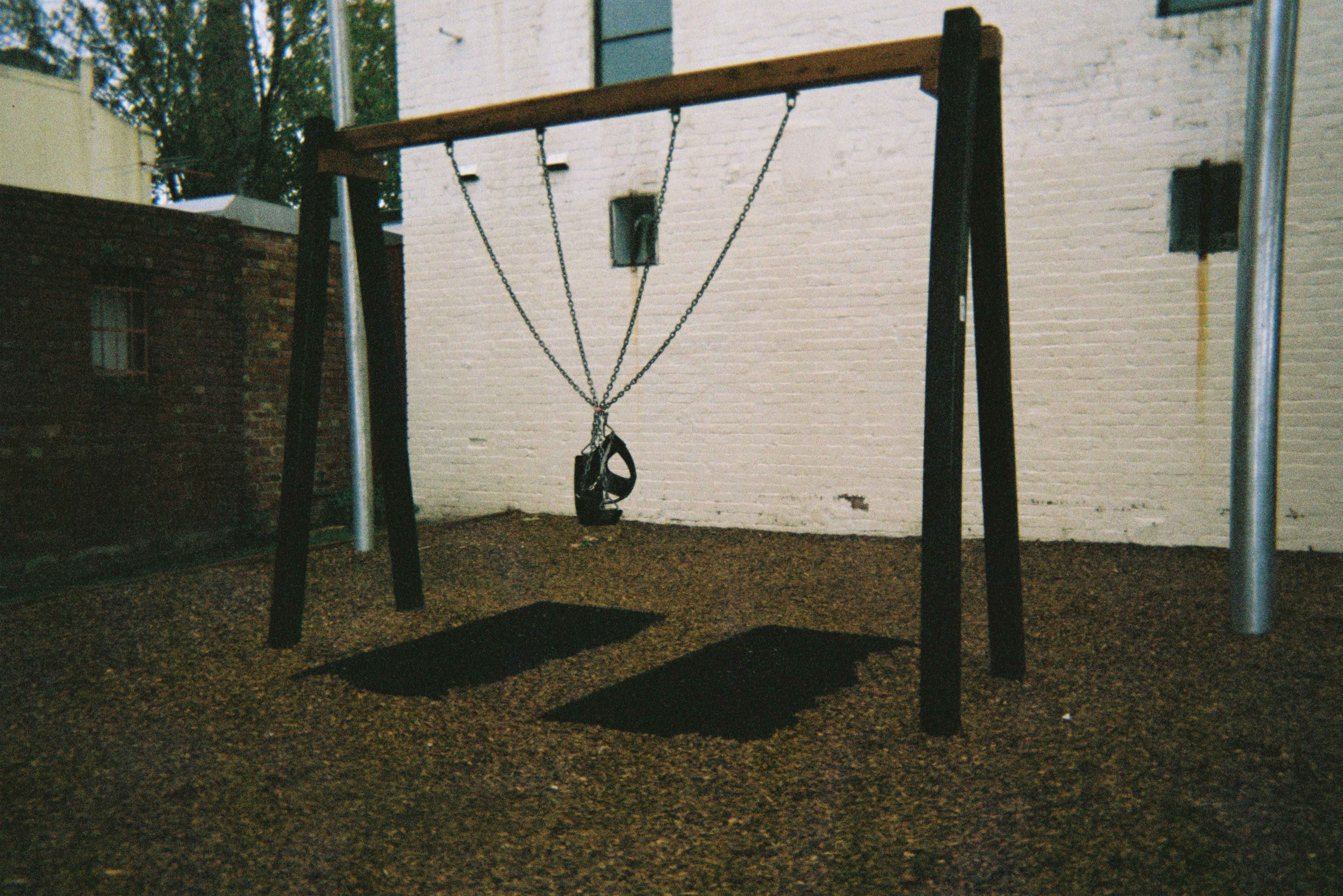 An empty swing set in a gravel playground, casting long shadows against a textured wall. The scene evokes nostalgia and stillness.