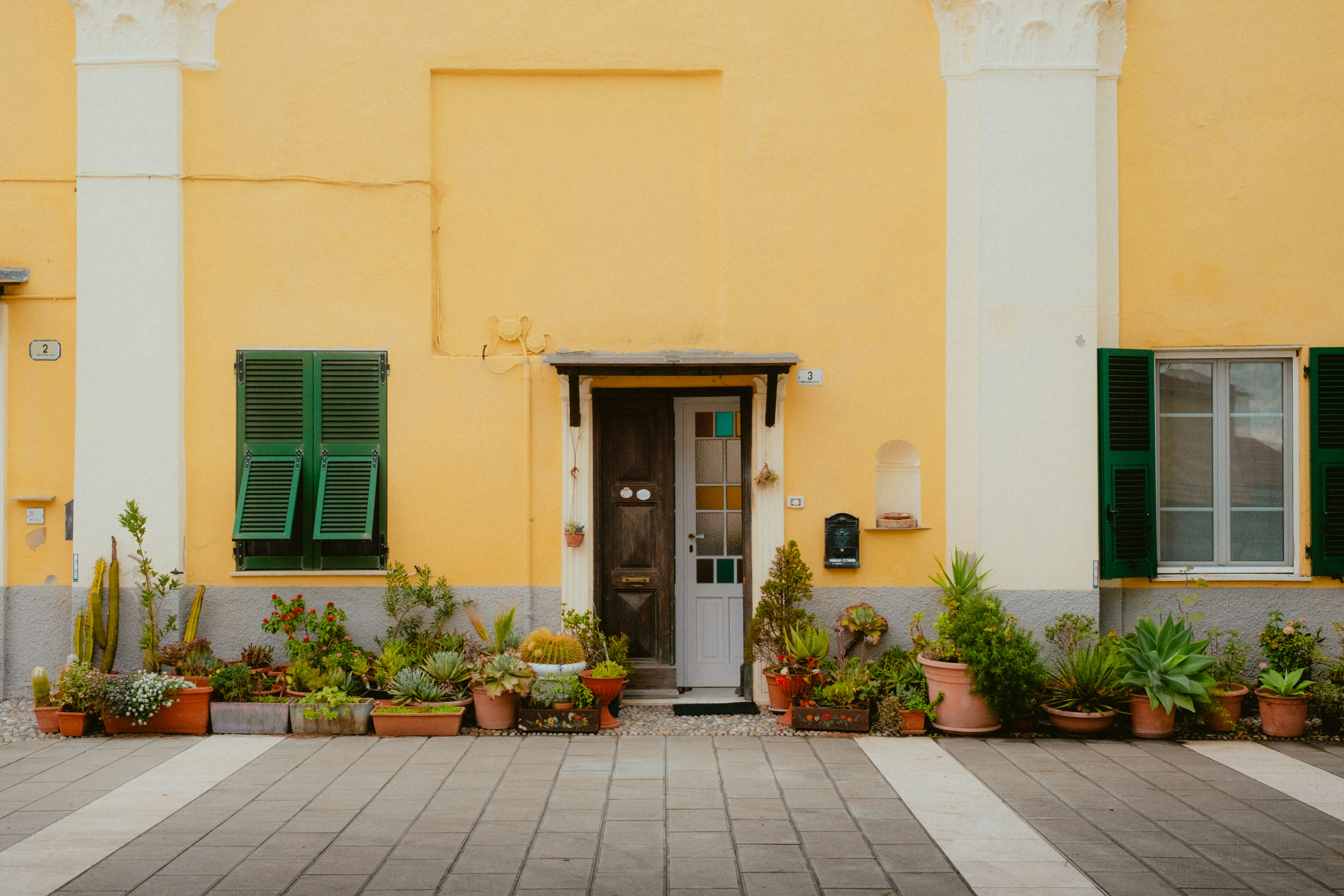 Yellow building with green shutters and a row of potted plants lining the entrance.