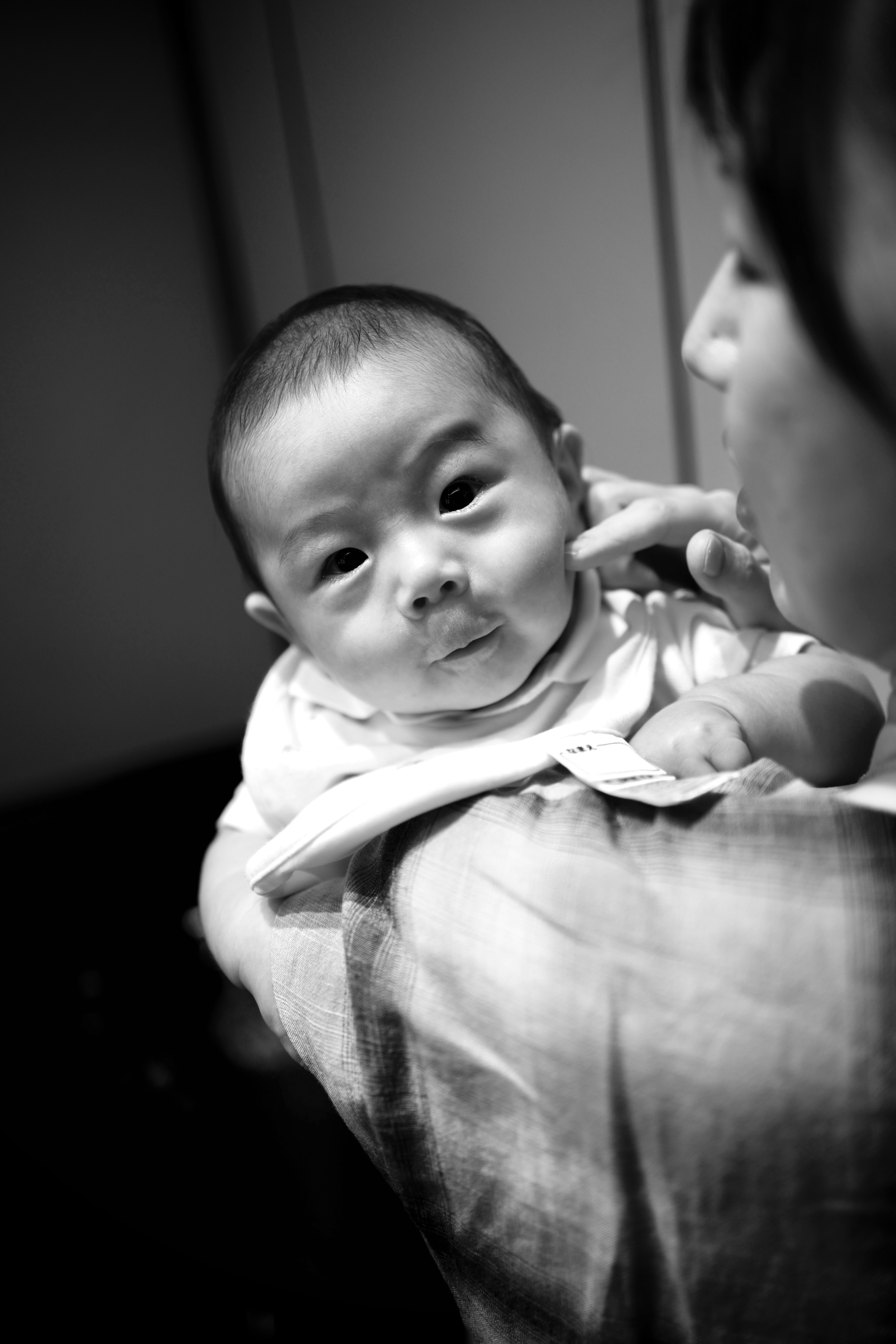 Infant gazing curiously while being held, with a gentle touch from an adult nearby. Black and white tones enhance the emotional depth.