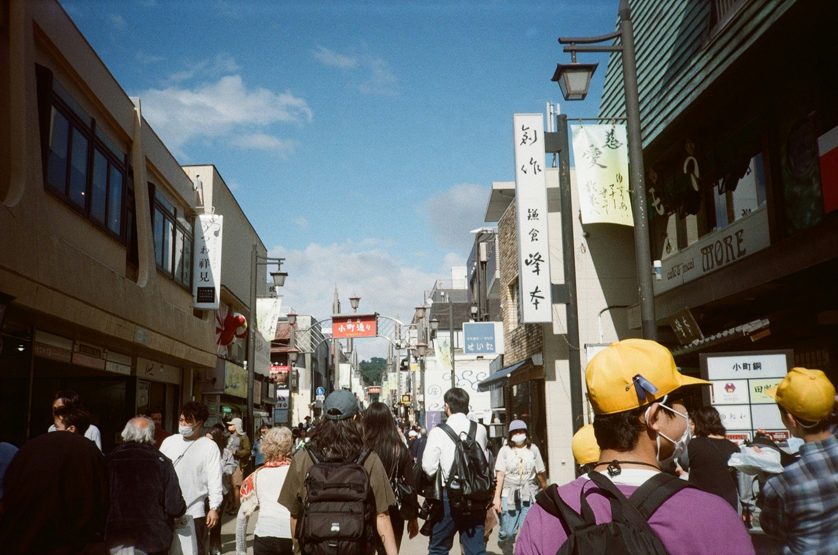 Crowded urban shopping street with storefronts, signs, and banners under a bright sky. A yellow-capped passerby anchors the foreground as pedestrians fill the scene.