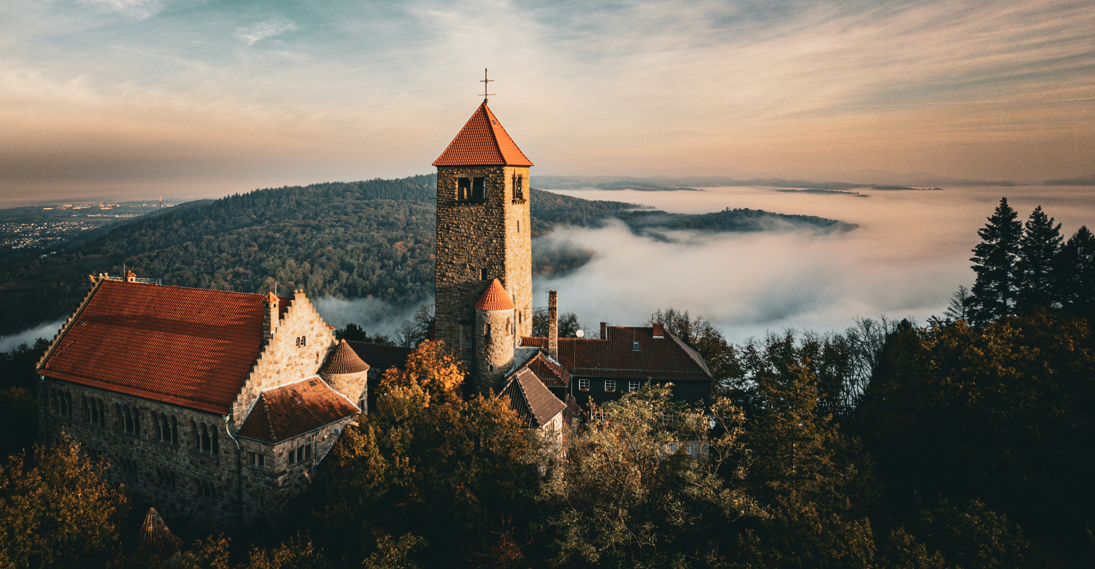 Aerial view of Wachenburg castle perched on a hilltop surrounded by autumn foliage and morning fog. The castle's stone tower and red-tiled roofs stand out against the misty landscape, with forested hills stretching into the distance. The soft light of sunrise creates a serene and atmospheric scene, highlighting the historic architecture of the castle amidst the tranquil natural surroundings.