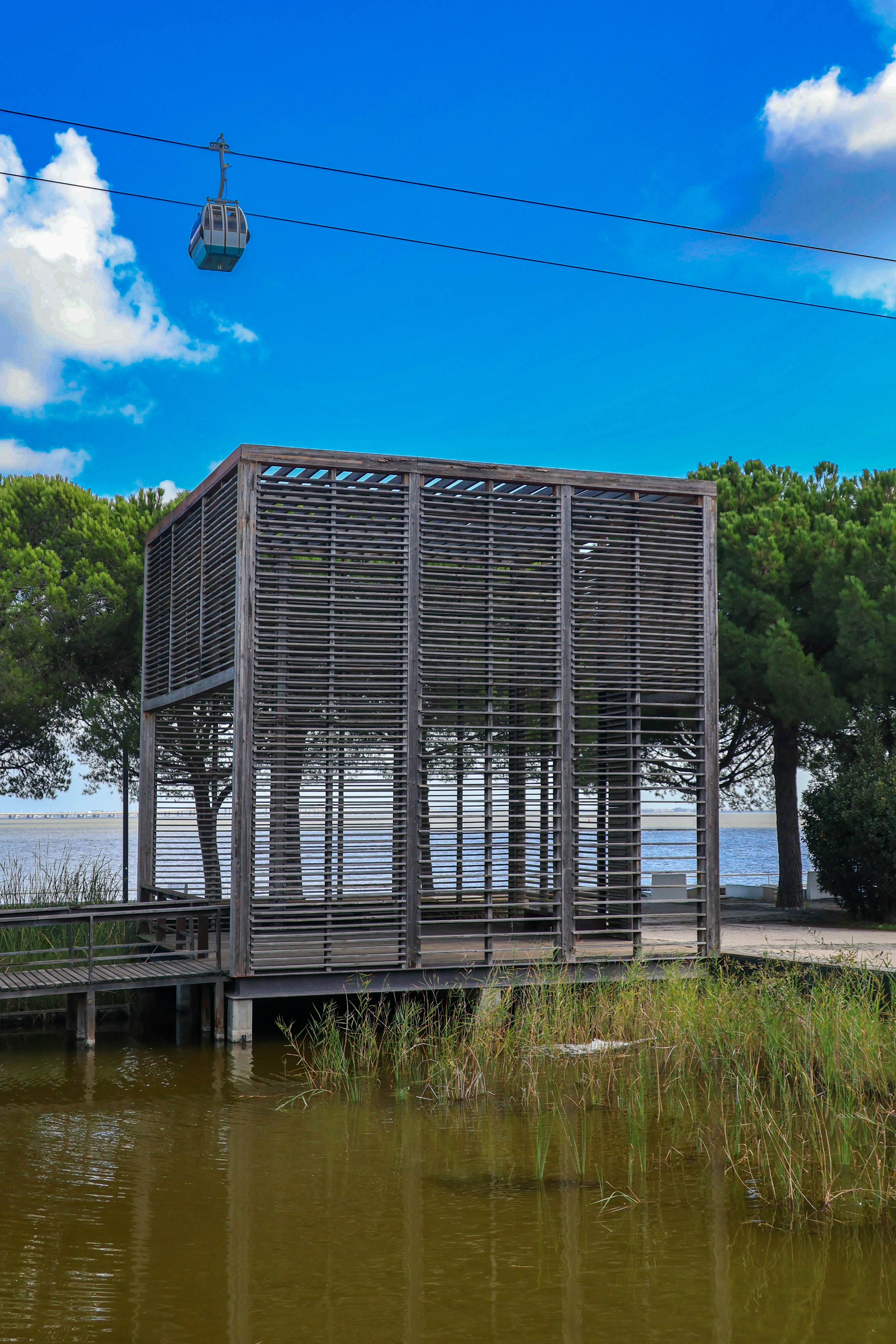 Lattice Pavilion by the Lake: Pines, Water, and a Gondola in the Bright Sky