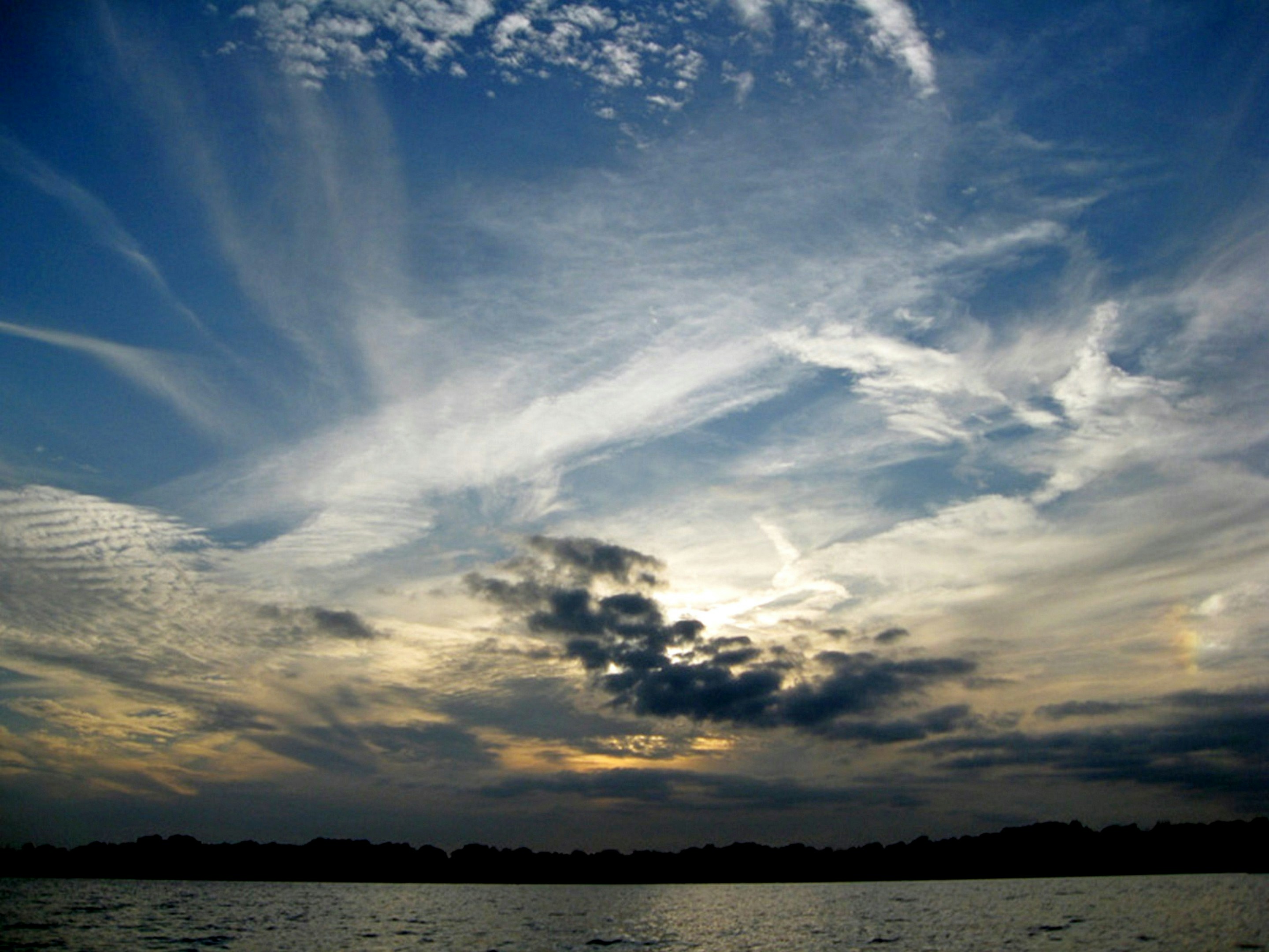 Dusk settles over a calm lake with dramatic cloud wisps sweeping across the sky. The expansive scene highlights warm tones and a subtle reflection on the water.