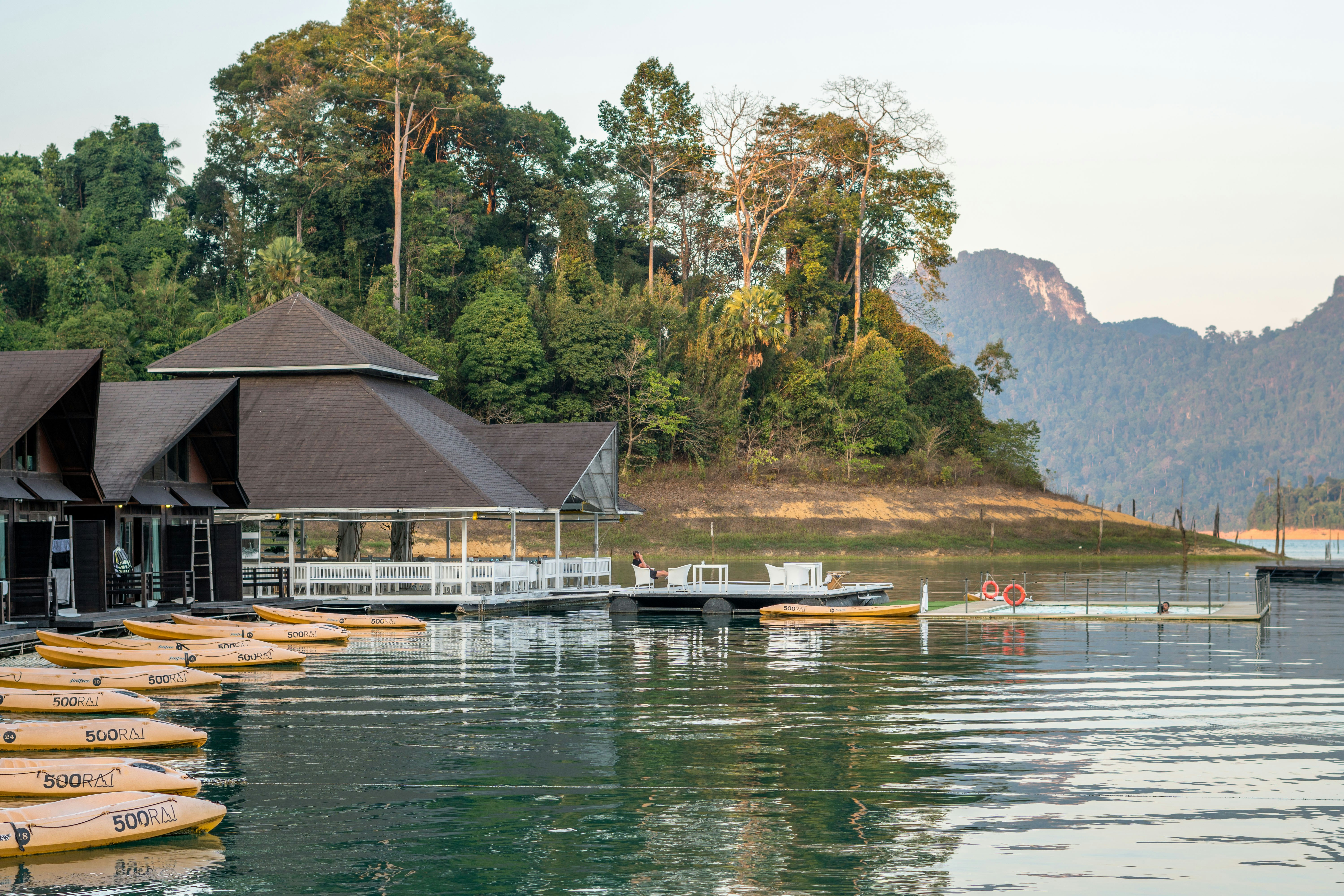 Boats lined up on a tranquil lake shore, with a wooden structure nestled among lush greenery and distant mountains. The calm waters reflect the surrounding scenery.