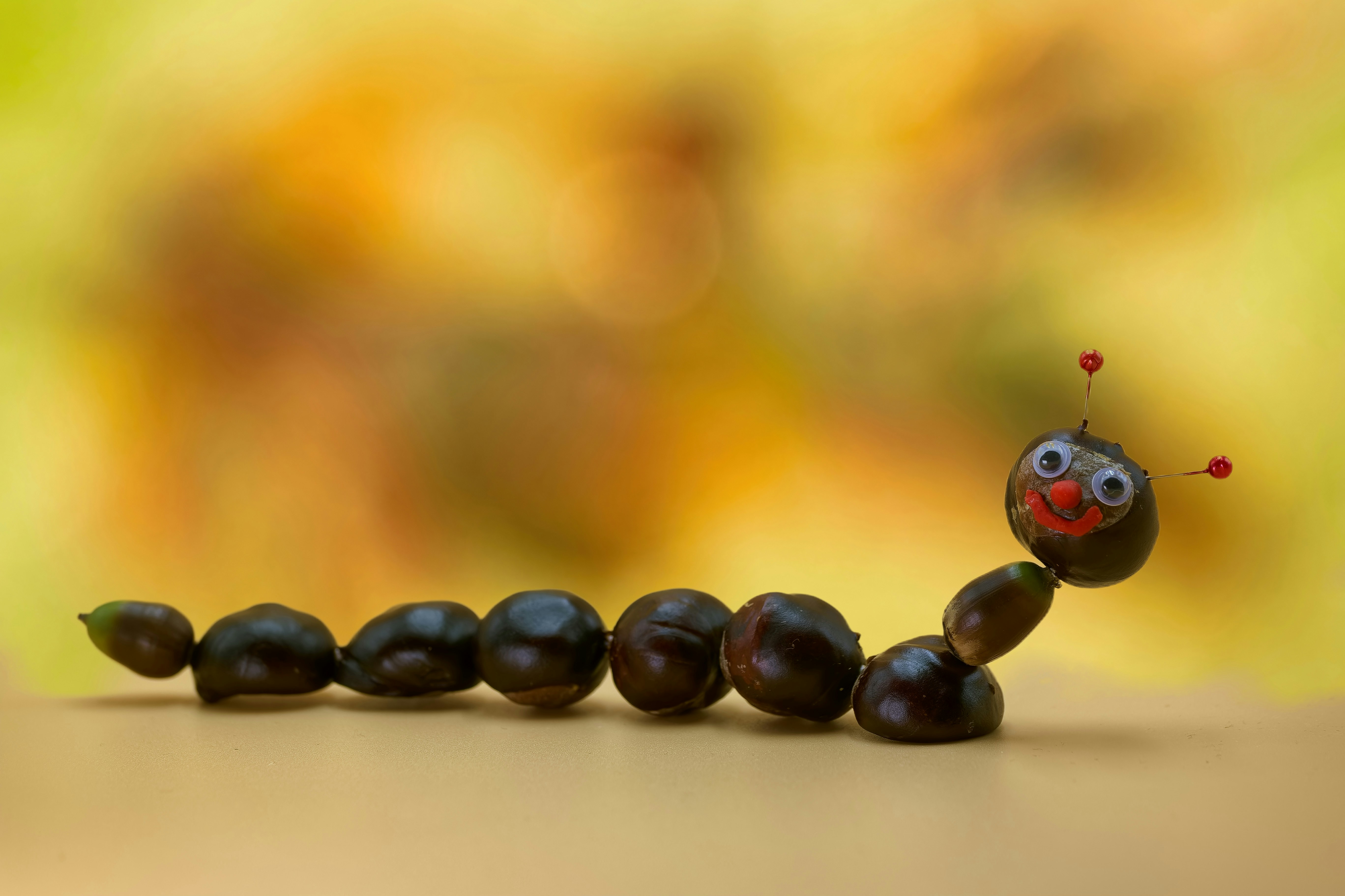 Macro photograph of glossy beans arranged in a line with a tiny bead-faced critter perched at the end, set against a warm, blurred background. A playful detail adds whimsy to the texture study.