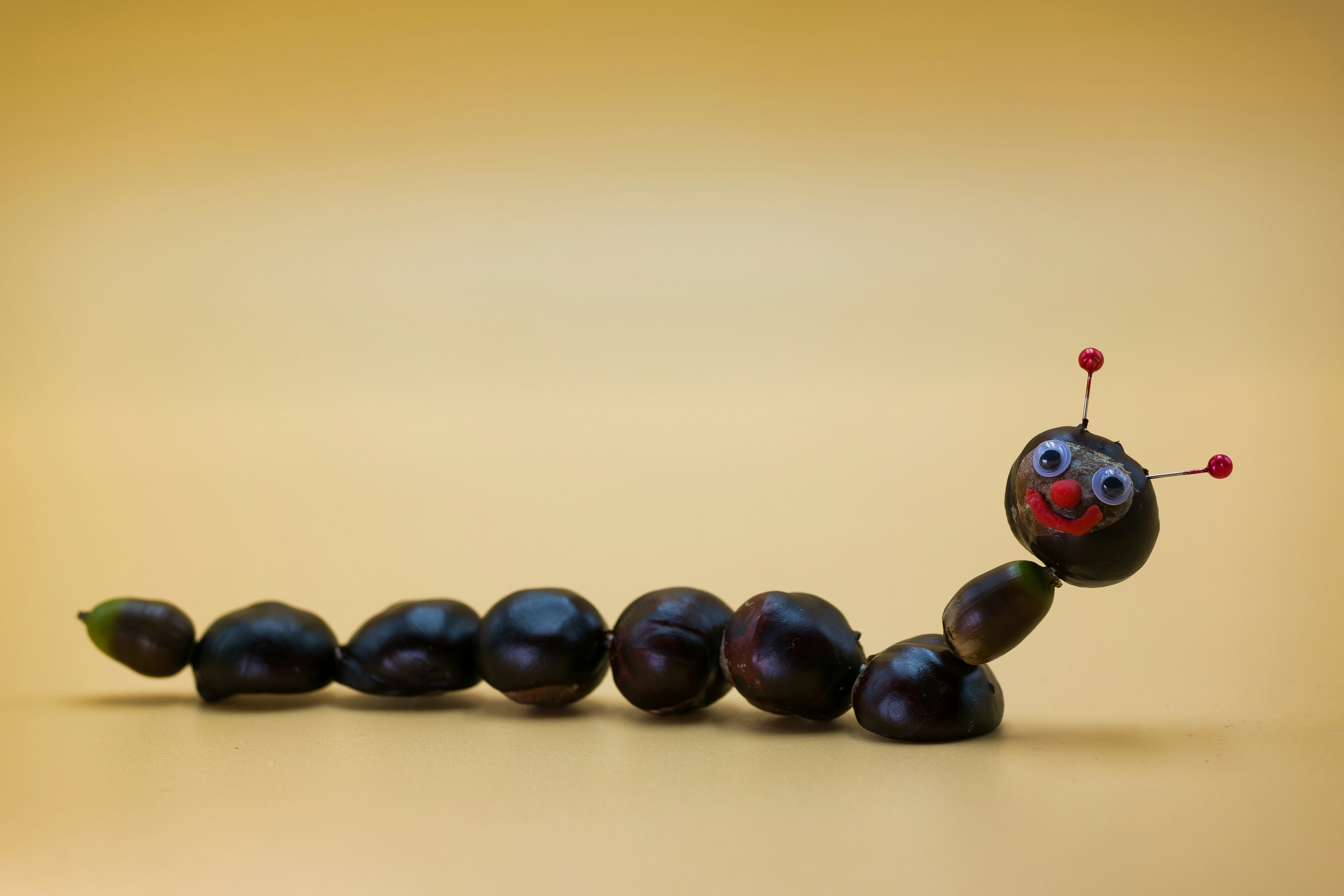 Row of glossy black beads arranged like an ant, with a small face on the final bead and a red antennae construct, set against a warm yellow background. A minimalist, toy-like composition emphasizing whimsy and color.