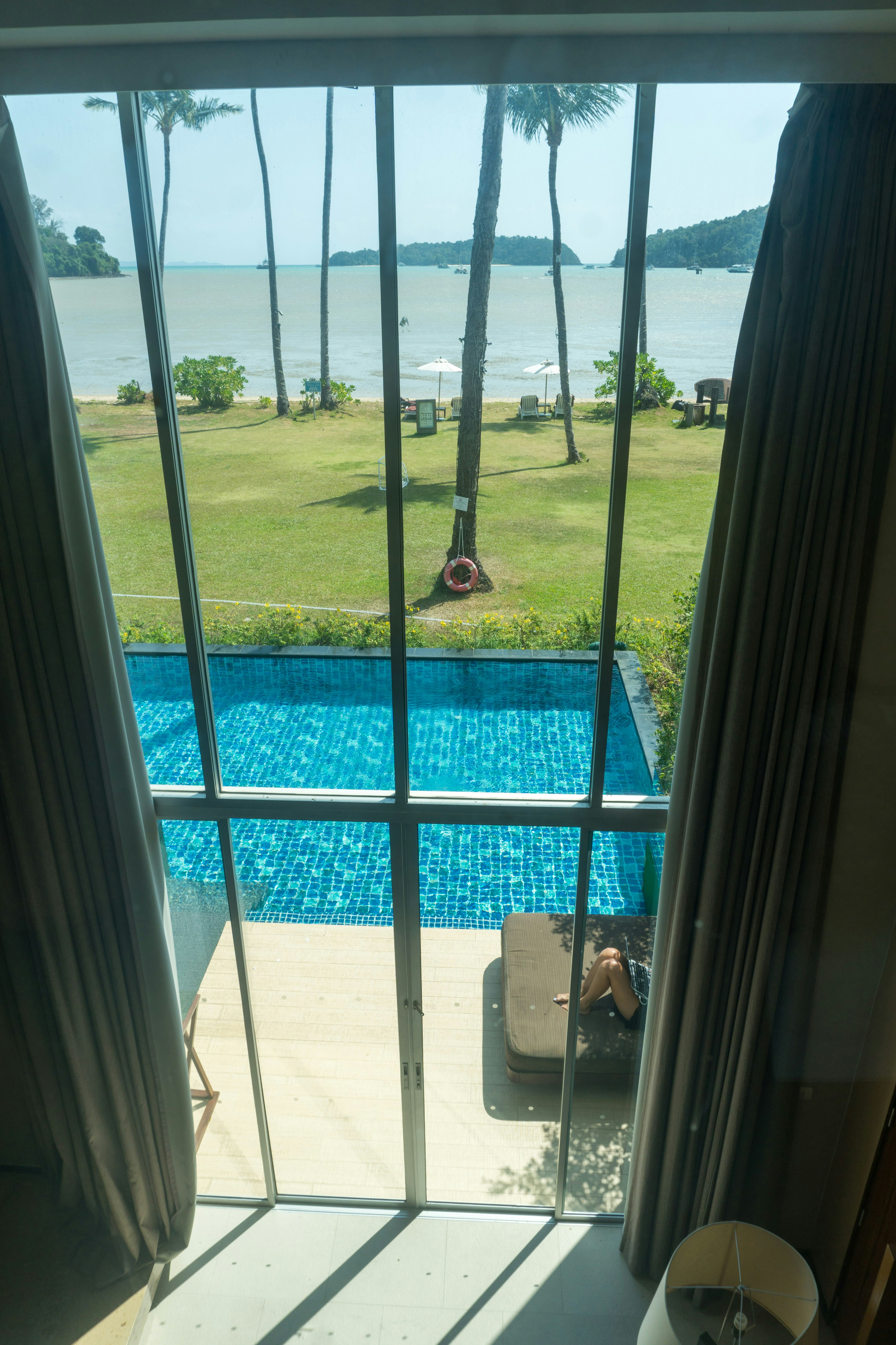 View from a room showcasing a poolside lounge chair, lush greenery, and a serene beach landscape beyond.