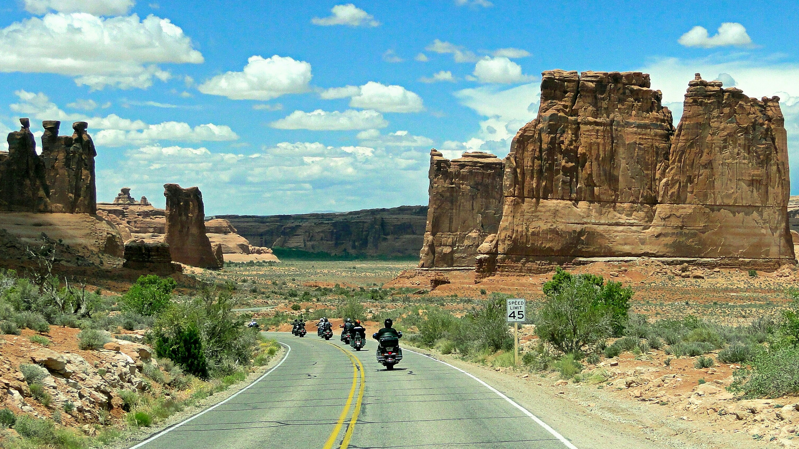 A winding desert highway threads between towering canyon spires as a small convoy of motorcyclists rides toward the horizon beneath a bright, cloud-speckled sky.