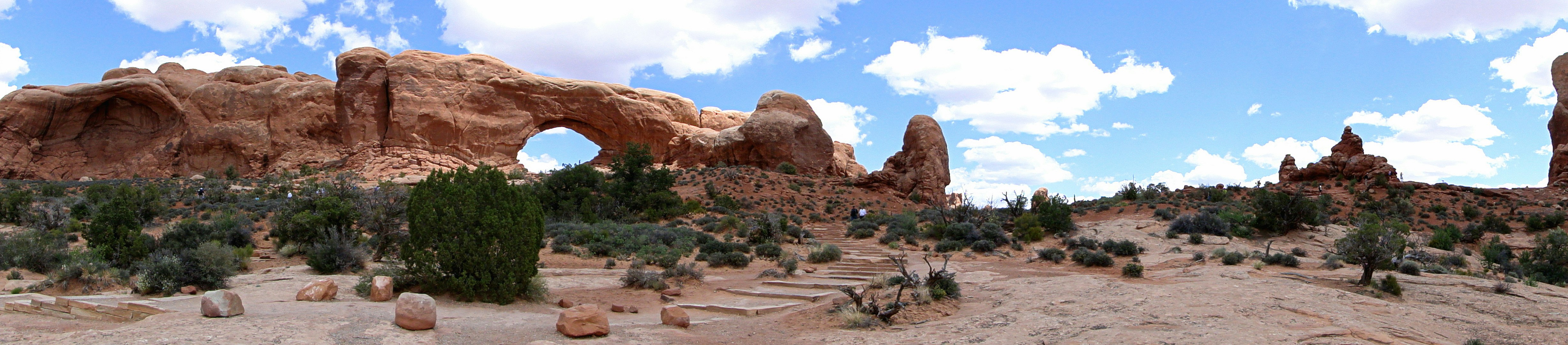 Panoramic desert landscape highlighting a prominent sandstone arch and scattered scrub beneath a bright, partly cloudy sky.
