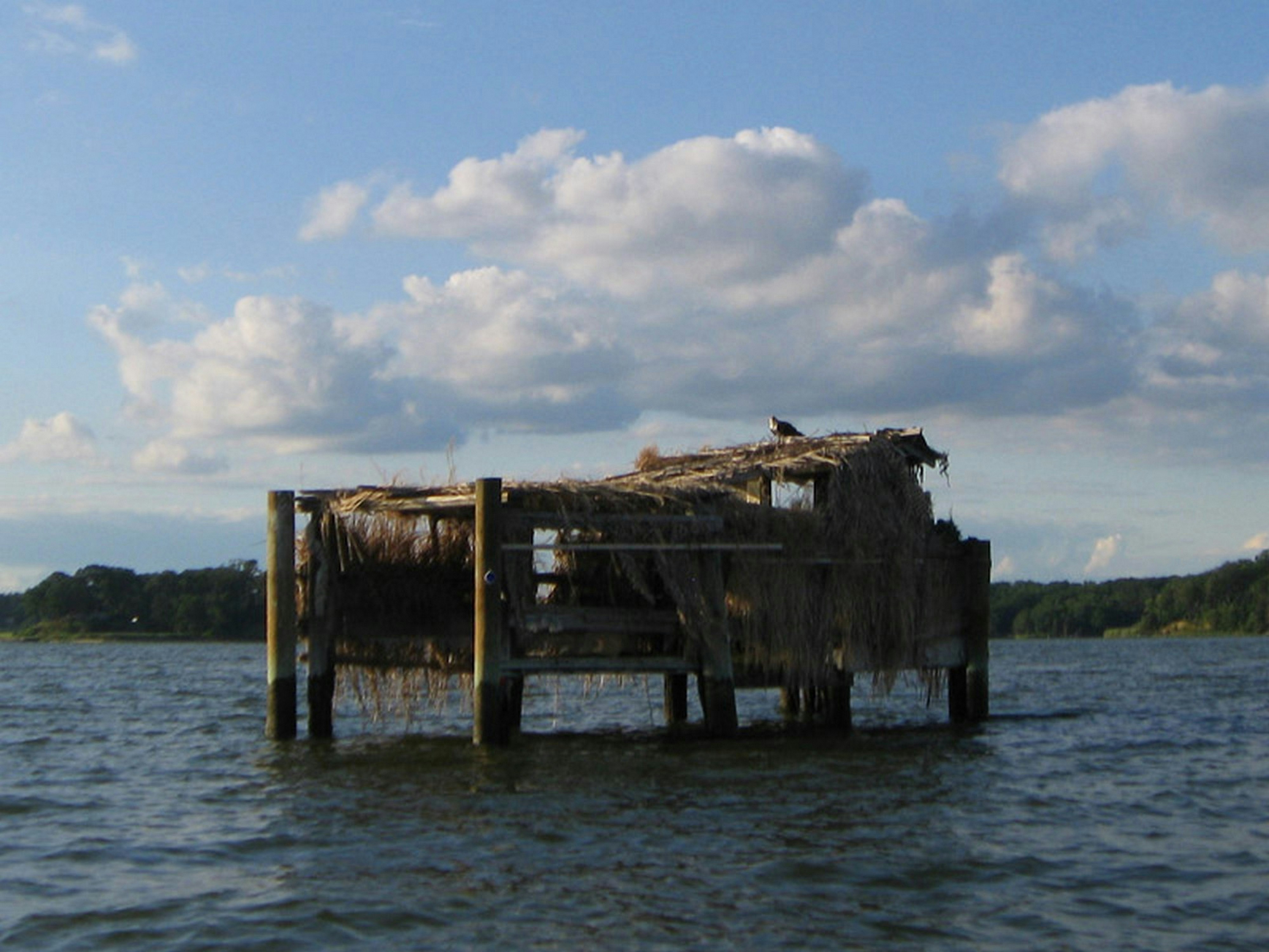 Thatched-roof wooden platform rises from a calm lake, supported by weathered pilings. Blue sky and distant shoreline emphasize the rustic, solitary structure.