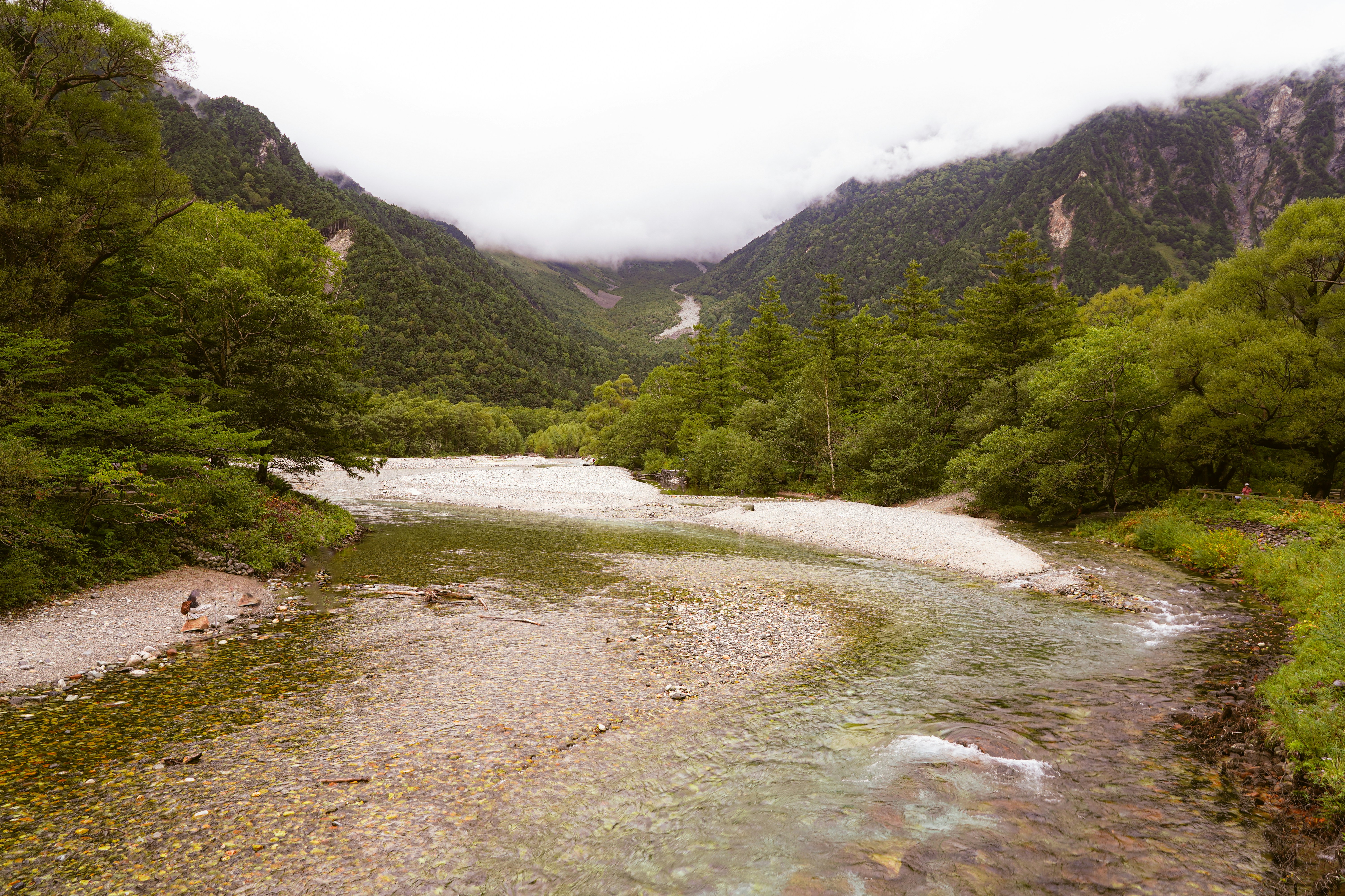 Meandering river flows through lush greenery and rocky banks, framed by towering mountains shrouded in mist.