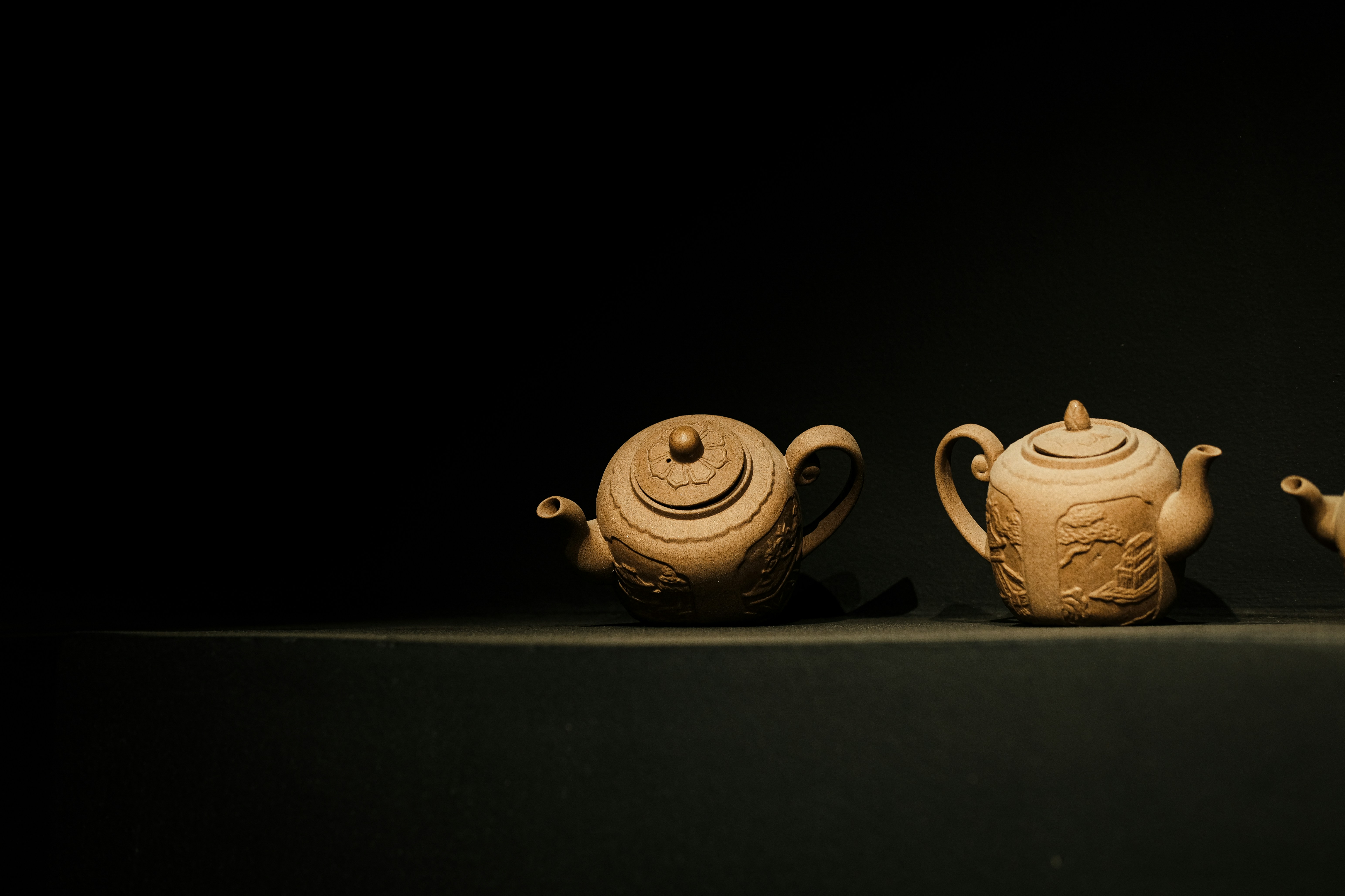 Clay teapots displayed on a dark shelf with intricate carvings highlighted by soft lighting.
