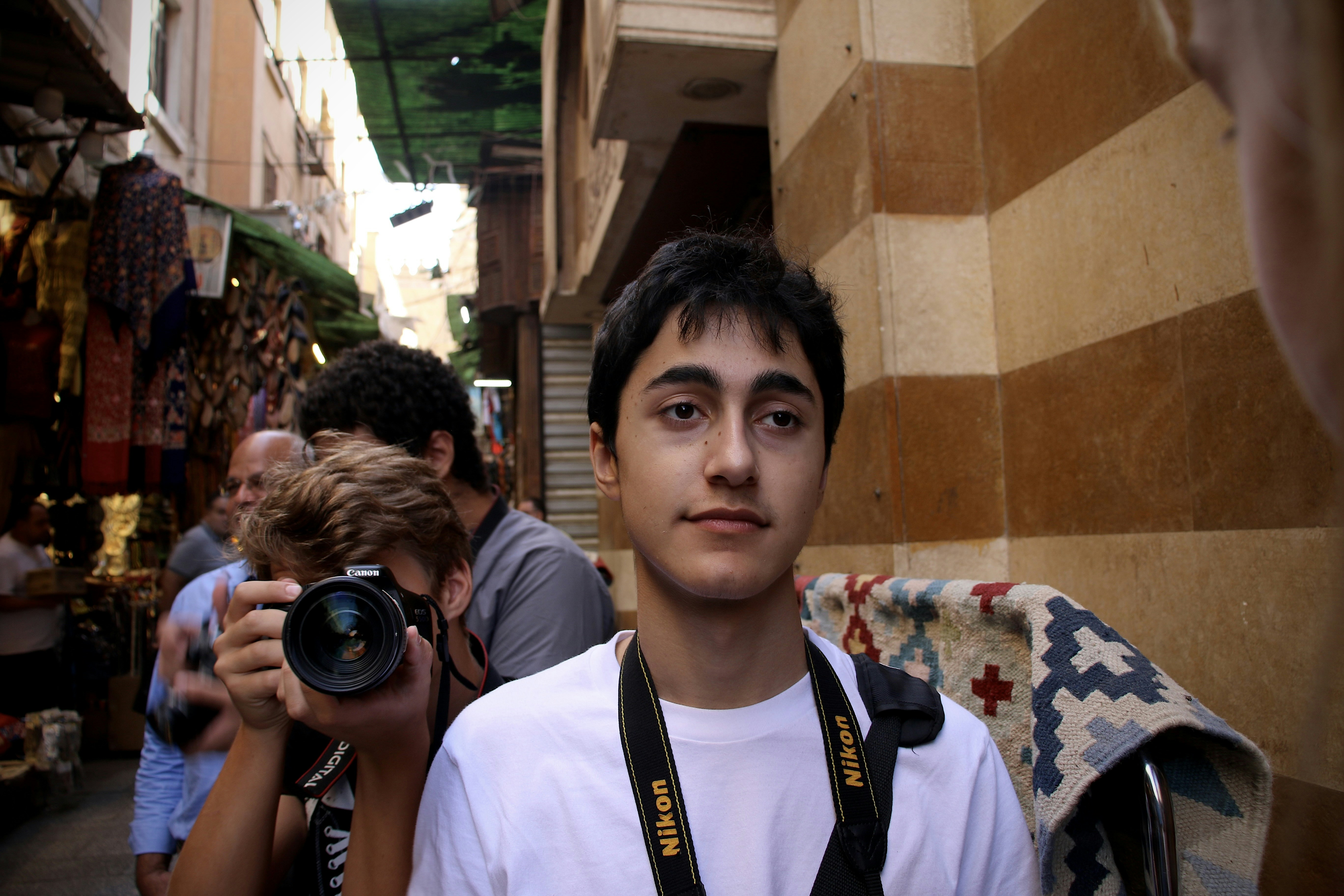 Young man with a camera strap stands in a bustling alley, surrounded by vibrant market activity.