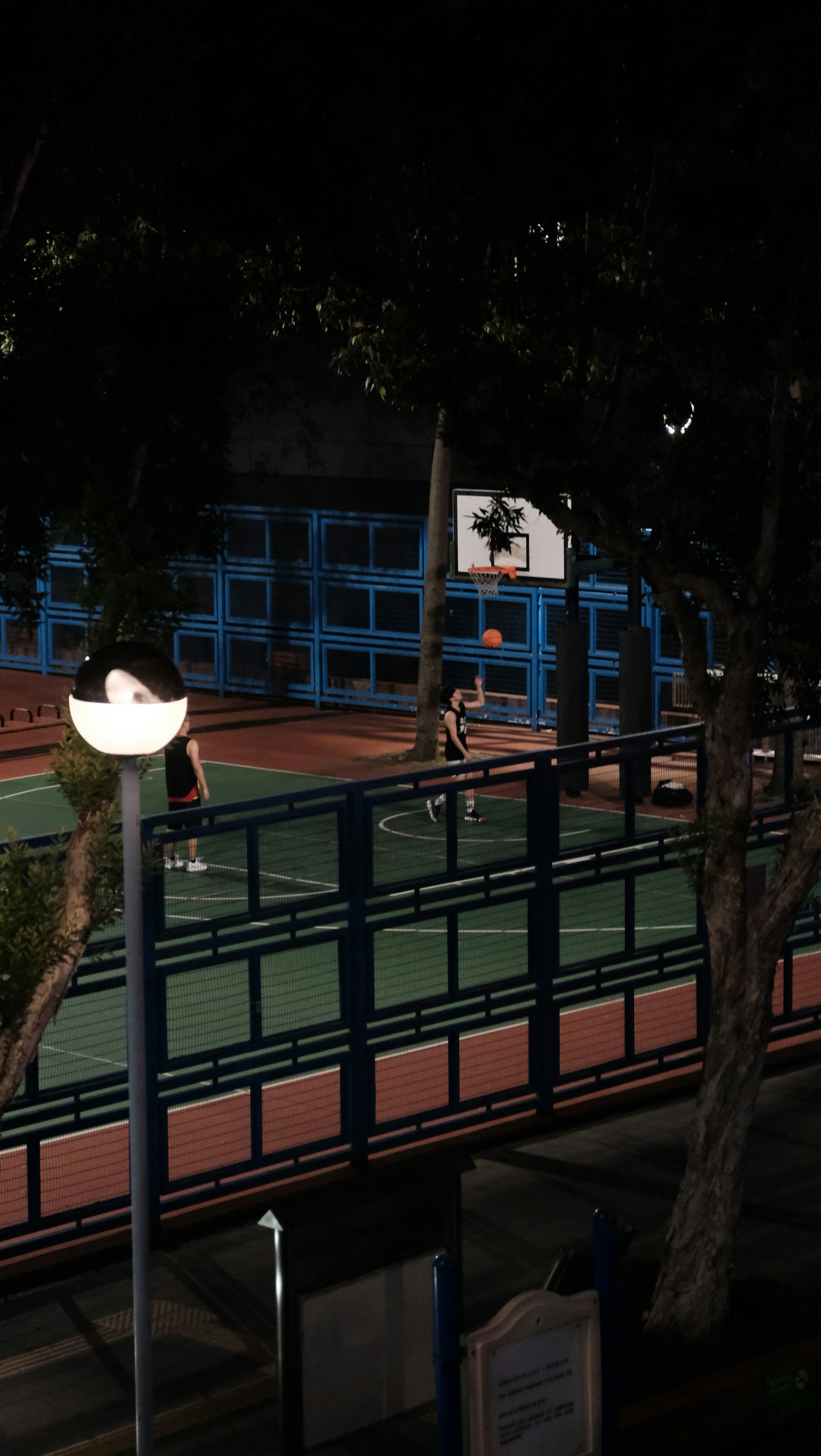 Basketball players engaged in a game on a court illuminated by streetlights, surrounded by trees at night.