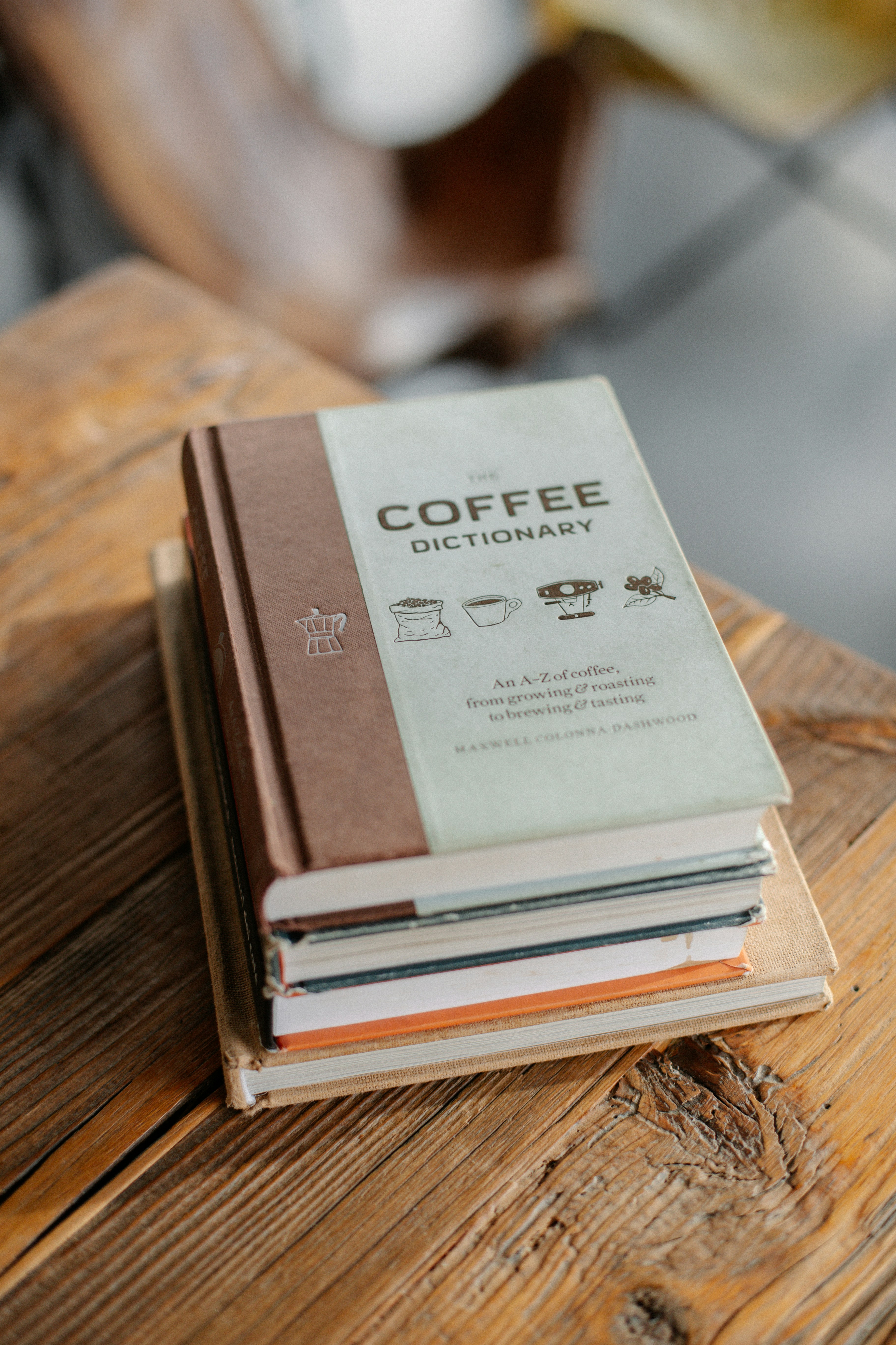 Stack of colorful books with 'The Coffee Dictionary' prominently displayed on top, resting on a wooden table. Perfect for coffee enthusiasts seeking to deepen their understanding.