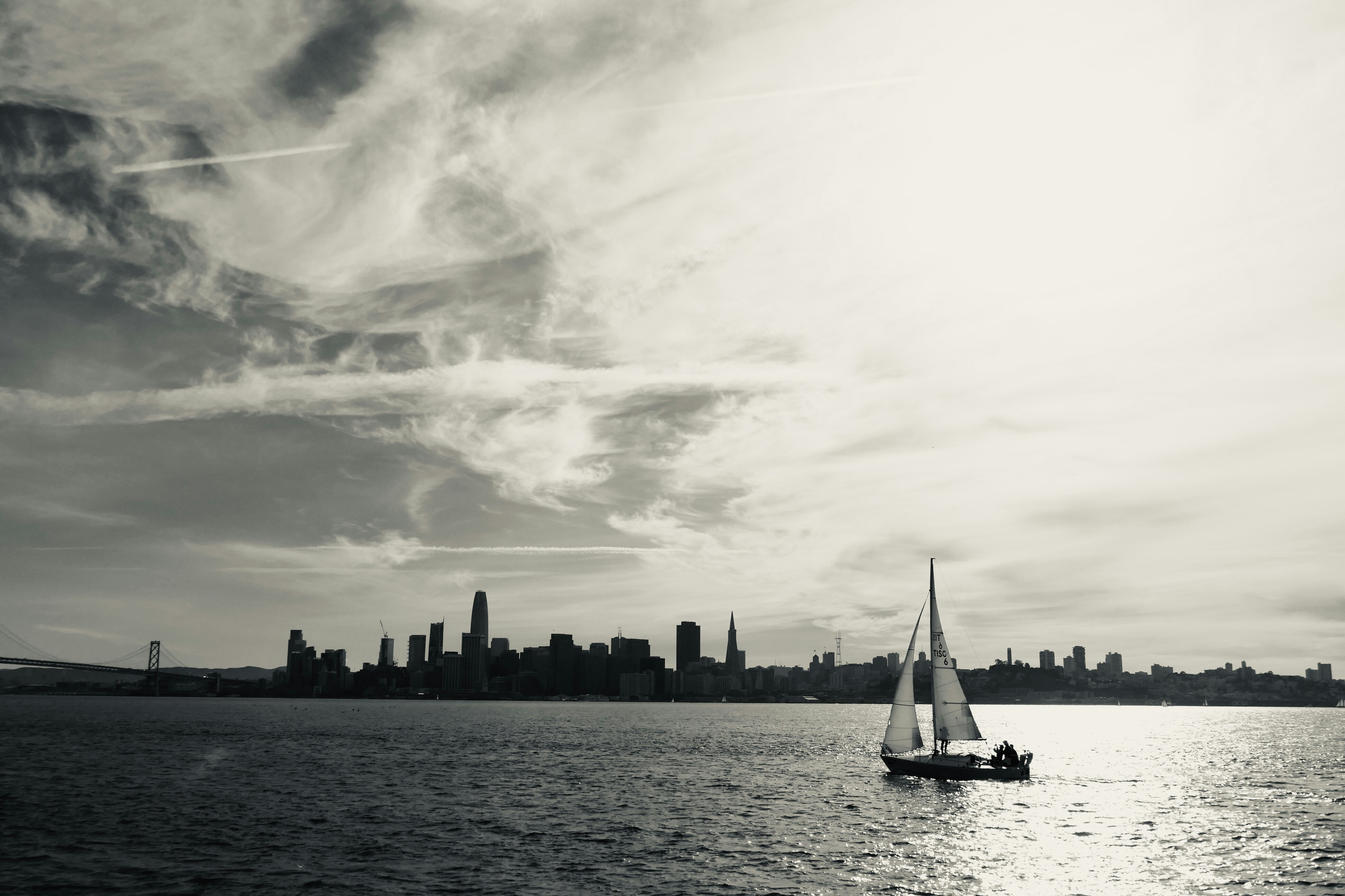 Sailboat gliding across shimmering waters with a silhouette of a city skyline under a dramatic sky. The scene evokes tranquility and adventure.