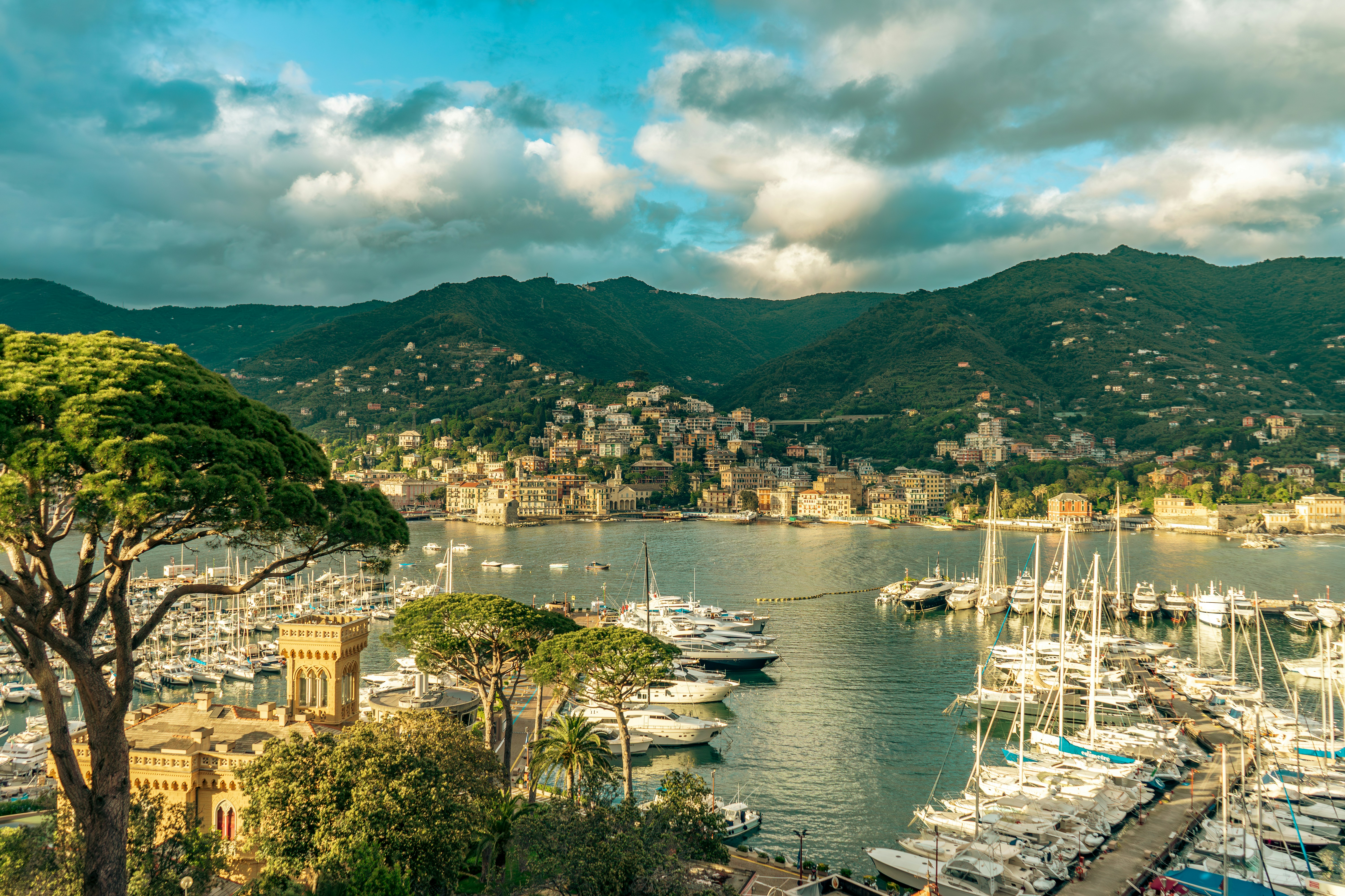 Boats anchored in a scenic harbor with lush hills and historic buildings in the background.