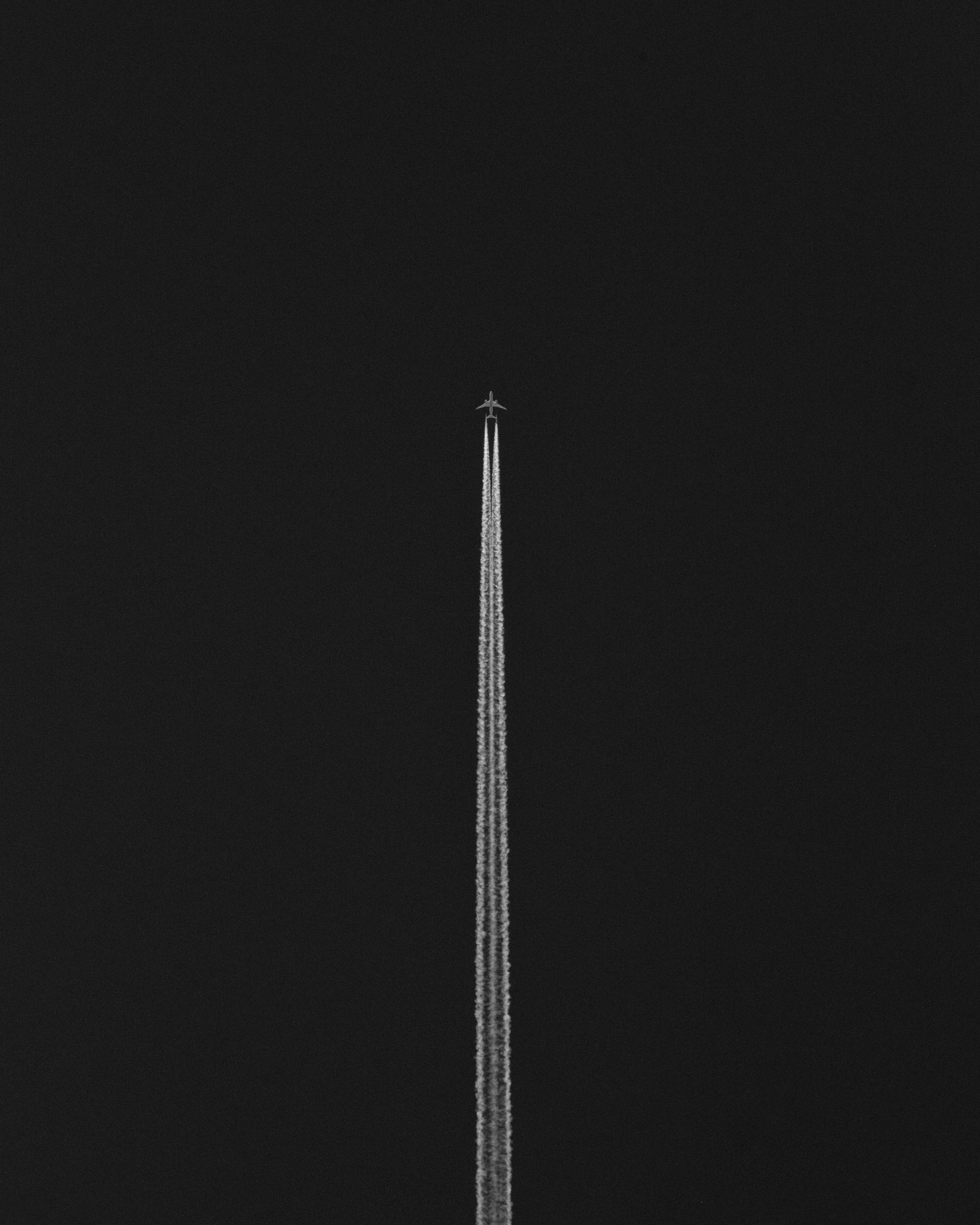 An airplane ascends against a stark black backdrop, leaving a thin trail of vapor in its wake.