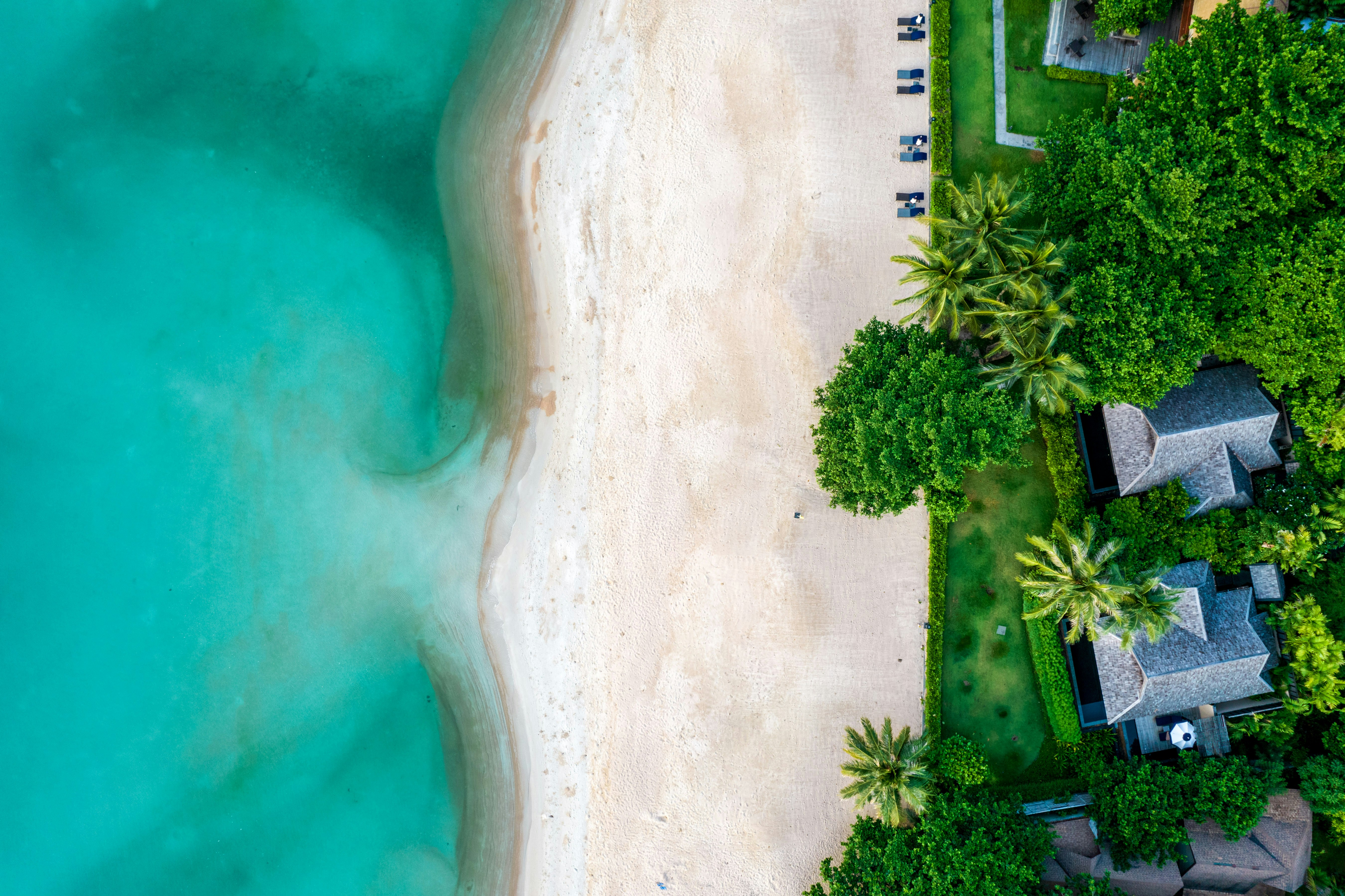 Aerial view of turquoise waters meeting a sandy beach lined with lush greenery and a nearby house.