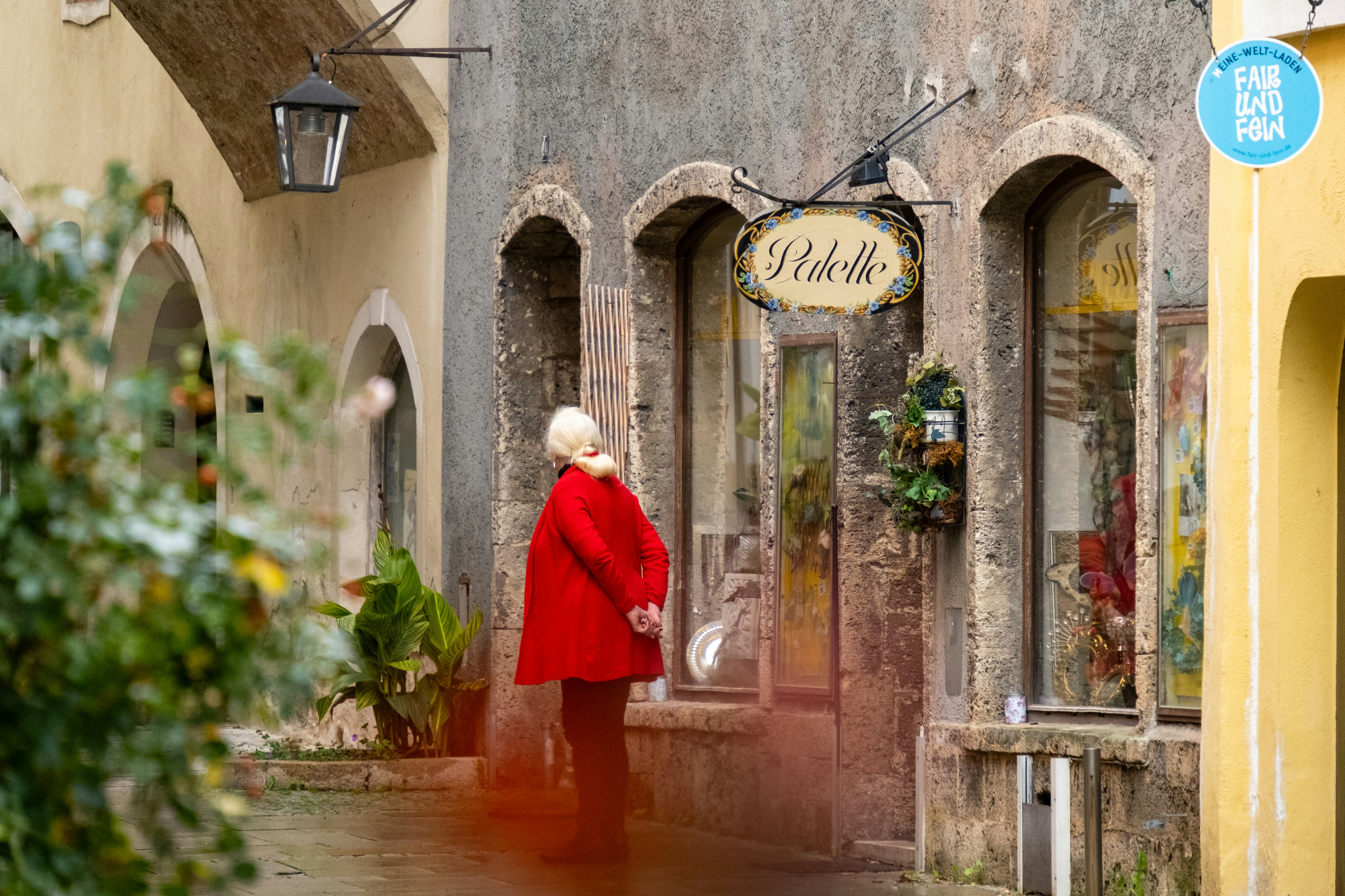 A man in a red coat stands before a rustic shop named "Palette" on a cobblestone street. Her back is to the camera as she gazes into the window, surrounded by charming old stone buildings with arched windows. Potted plants add greenery, and the colorful shop sign adds warmth. The red coat pops against the muted tones, creating a peaceful scene.