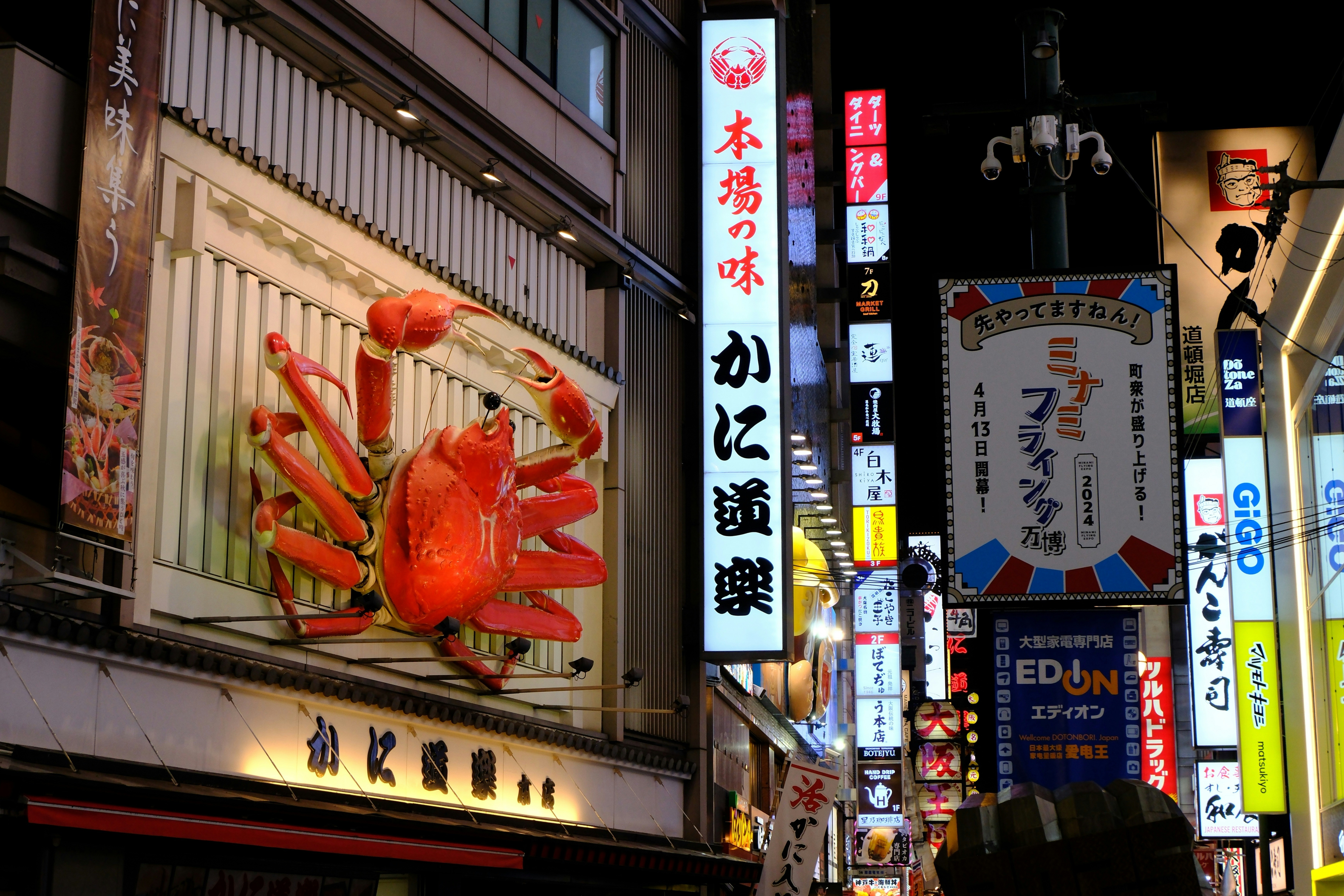 A photograph of a neon-lit street scene featuring a giant red crab sign above a restaurant, flanked by tall vertical Japanese signs.