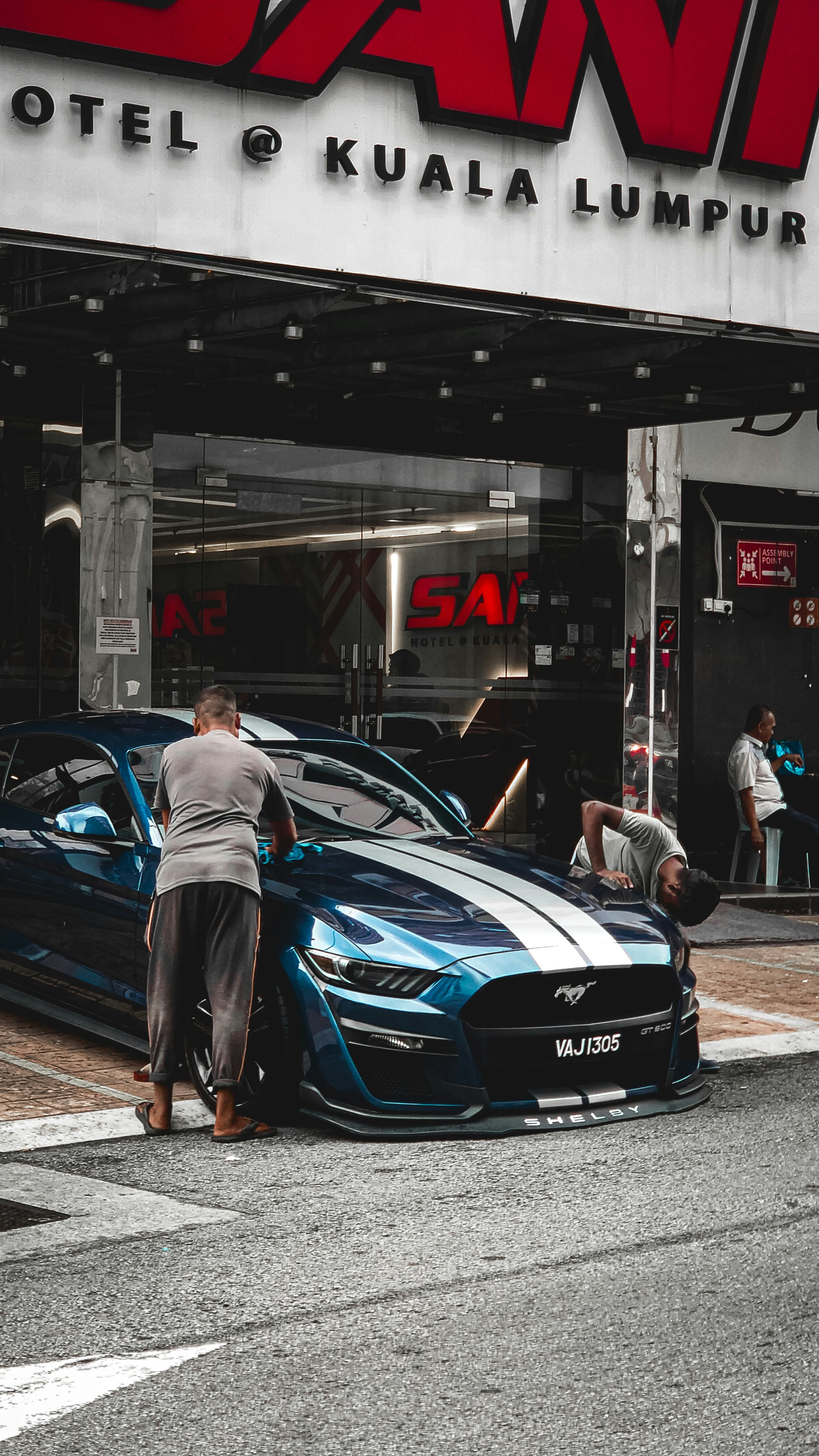 Blue Ford Mustang GT with white racing stripes parked outside a Kuala Lumpur hotel while two men work on the car.