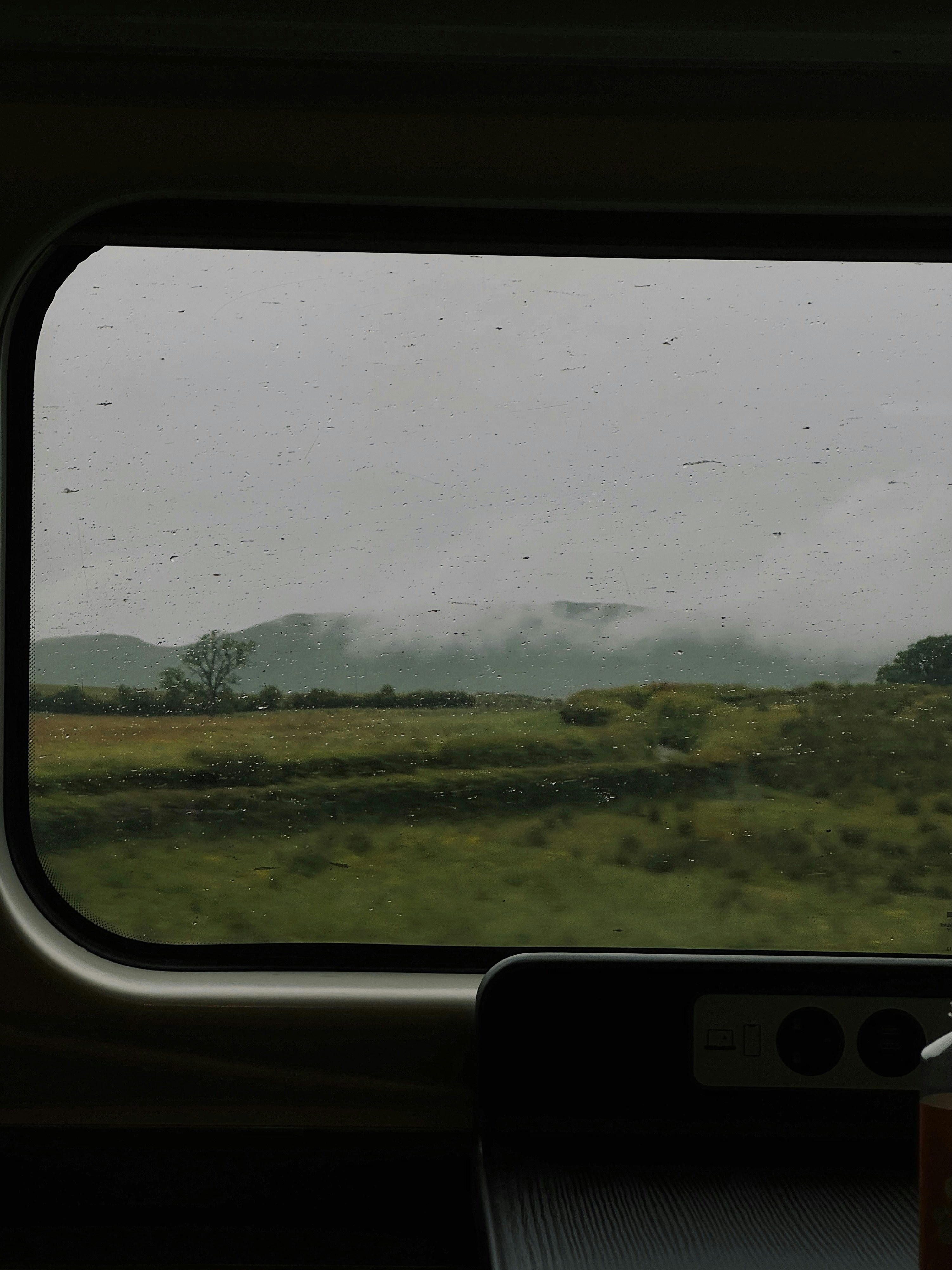 Rain-dusted window frames a misty landscape of fields and distant hills beneath an overcast sky.