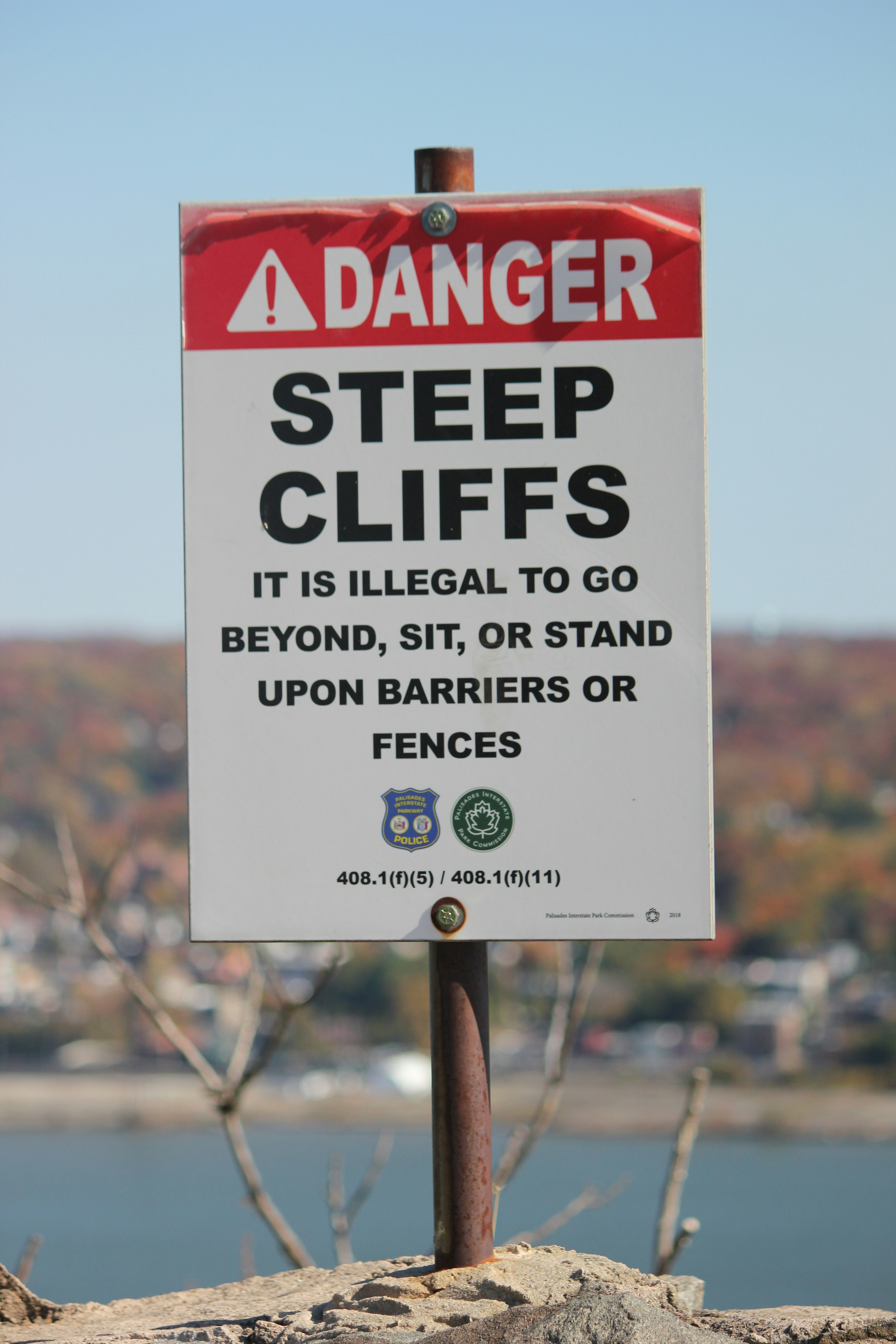 Sign warning of steep cliffs and legal restrictions against approaching barriers or fences. The sign is prominently displayed against a scenic backdrop.