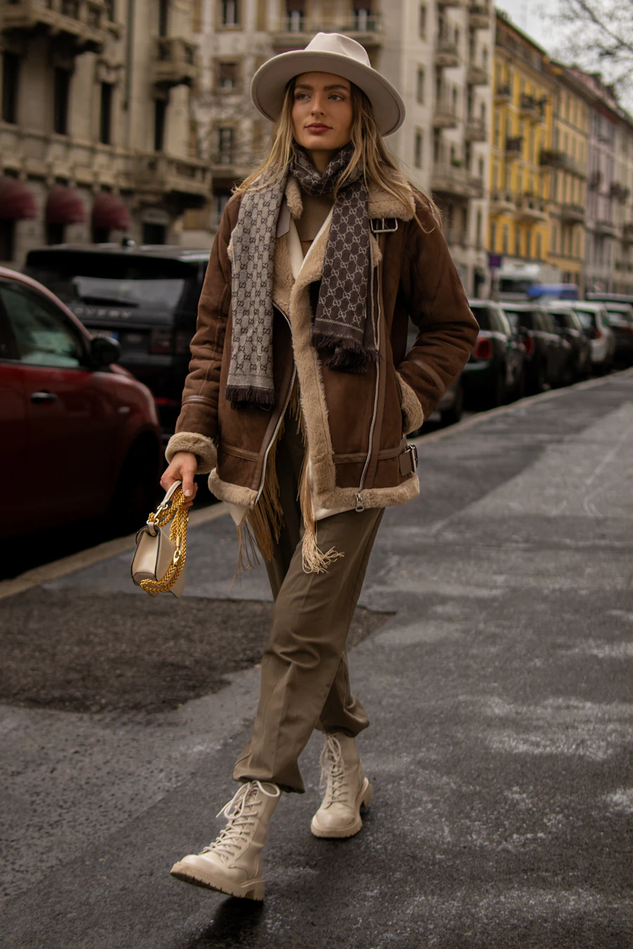 A woman walks down a city street in a brown shearling jacket, patterned scarf, wide-brim hat, and lace-up boots, carrying a small handbag. Parked cars and tall buildings line the background.